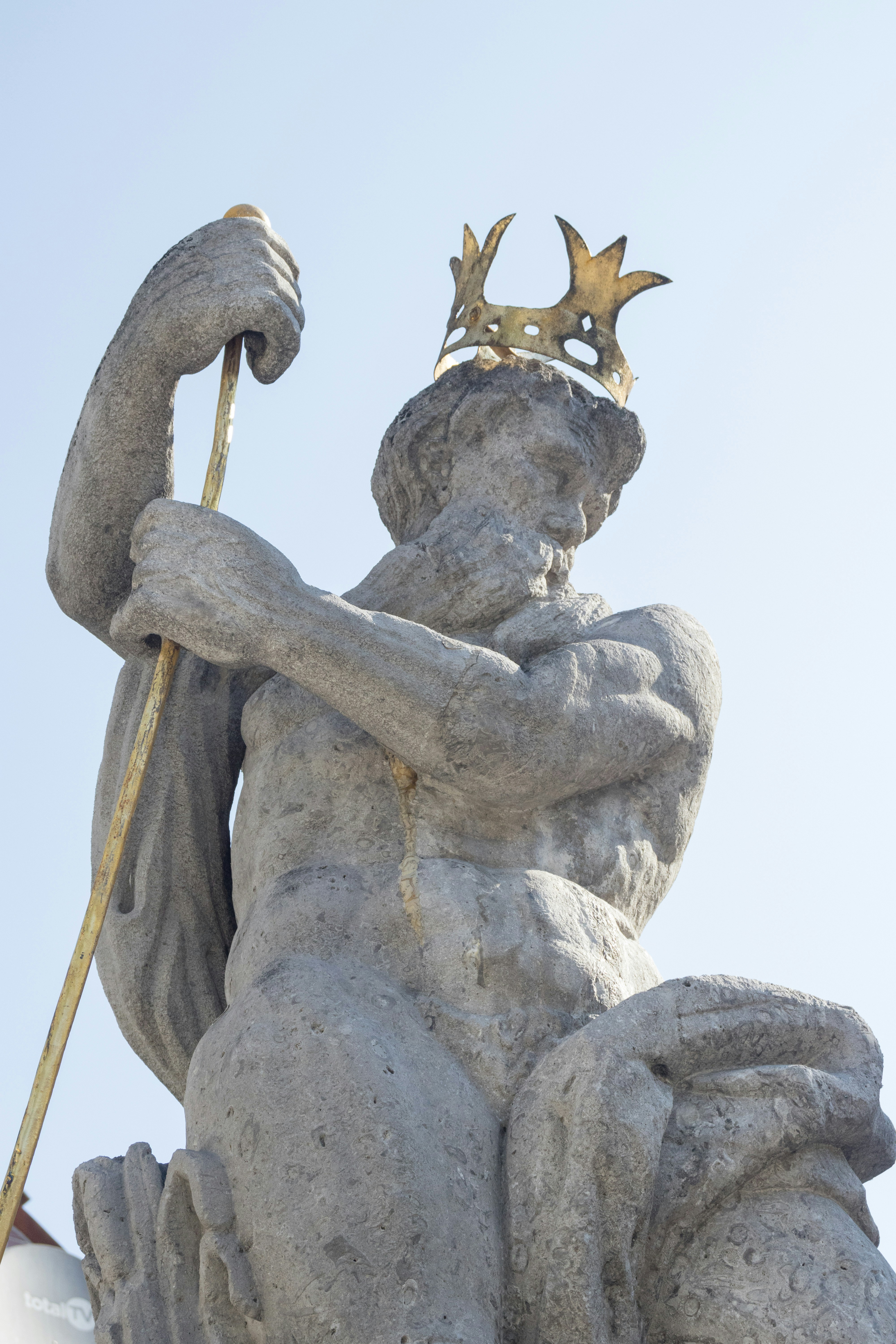 Stone statue holding a staff, wearing a regal crown, set against a clear blue sky. Close-up reveals weathered texture and sculpture details.