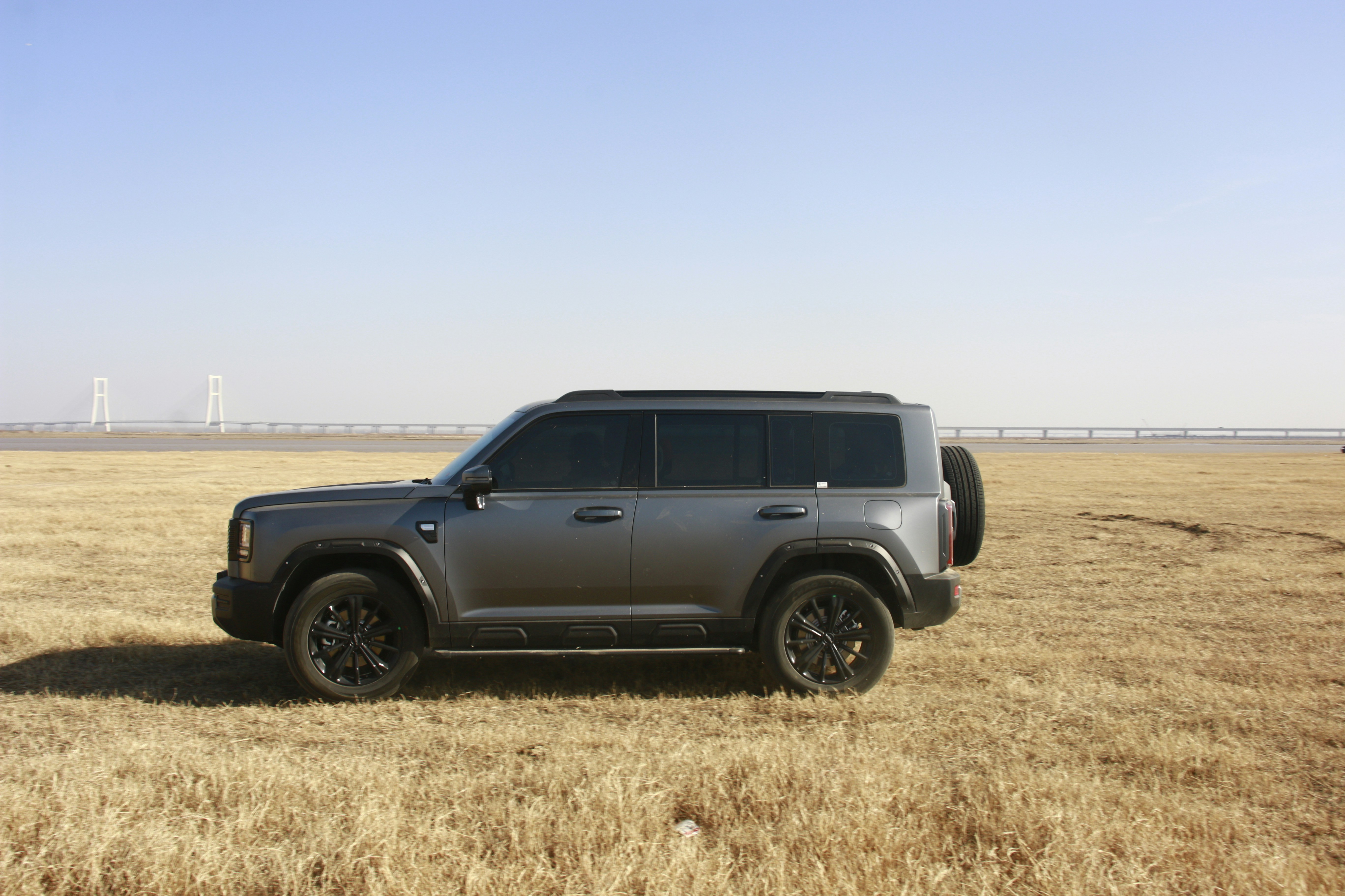 Grey SUV parked on dry field with a distant bridge on the horizon; a minimal landscape emphasizing the vehicle. This is a photographic shot of a real car in an open plain.