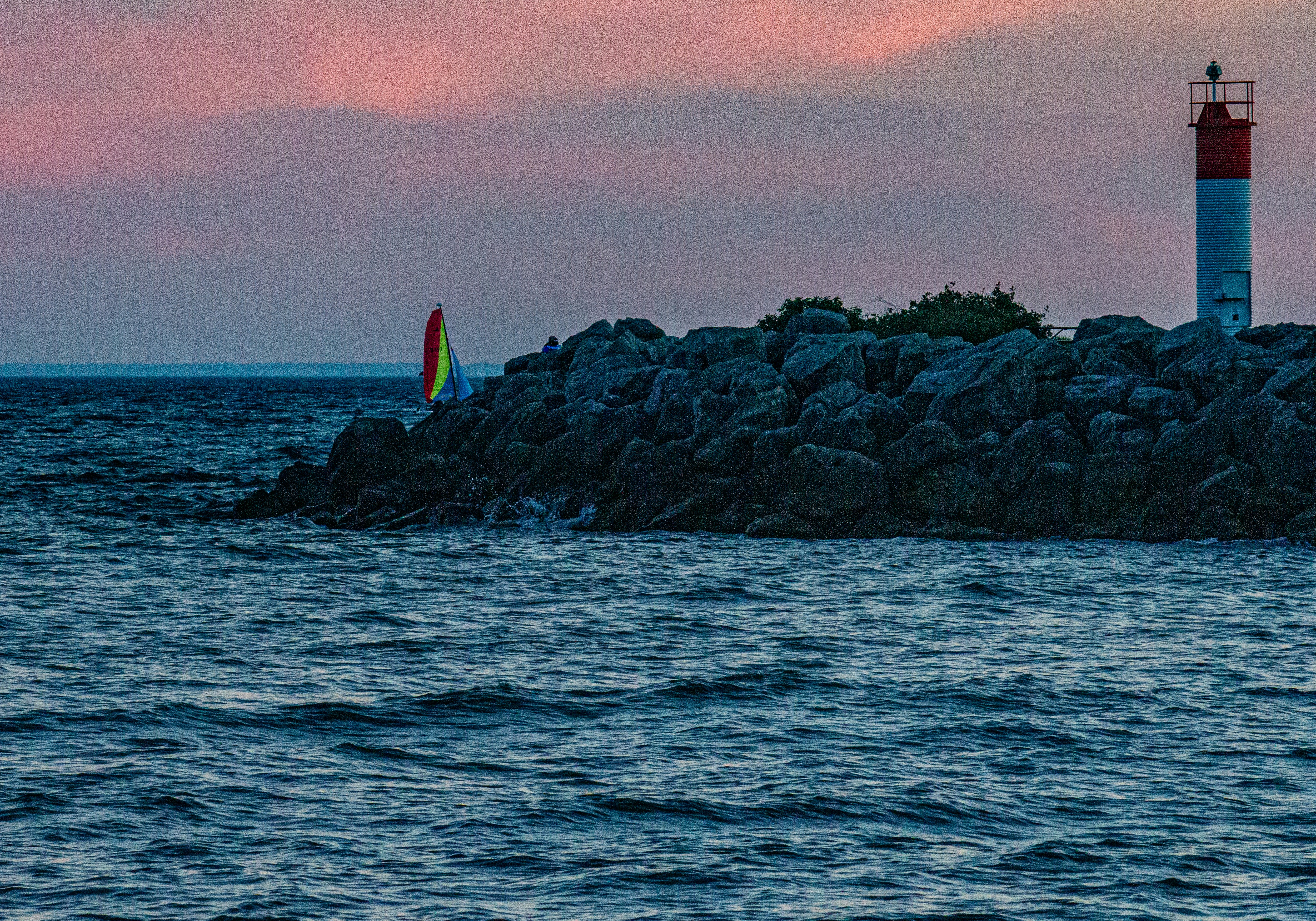 Lighthouse perched on rocky breakwater with a sailboat glancing by as evening light settles over calm sea.