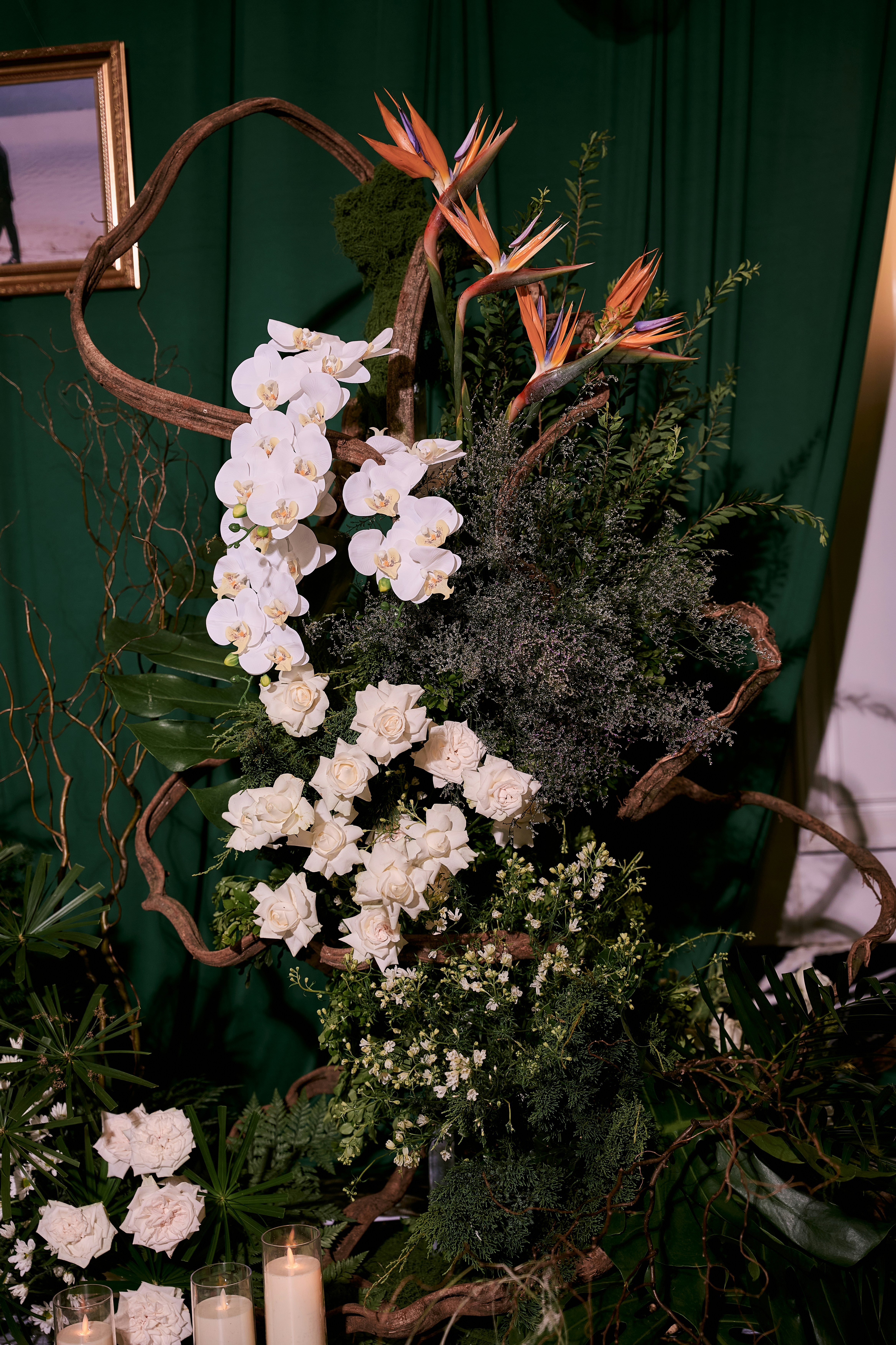 A lush floral display with white orchids and birds of paradise set against a deep green backdrop, accented by sculptural wood elements and candles. The arrangement dominates the frame as the centerpiece of a decorative installation.
