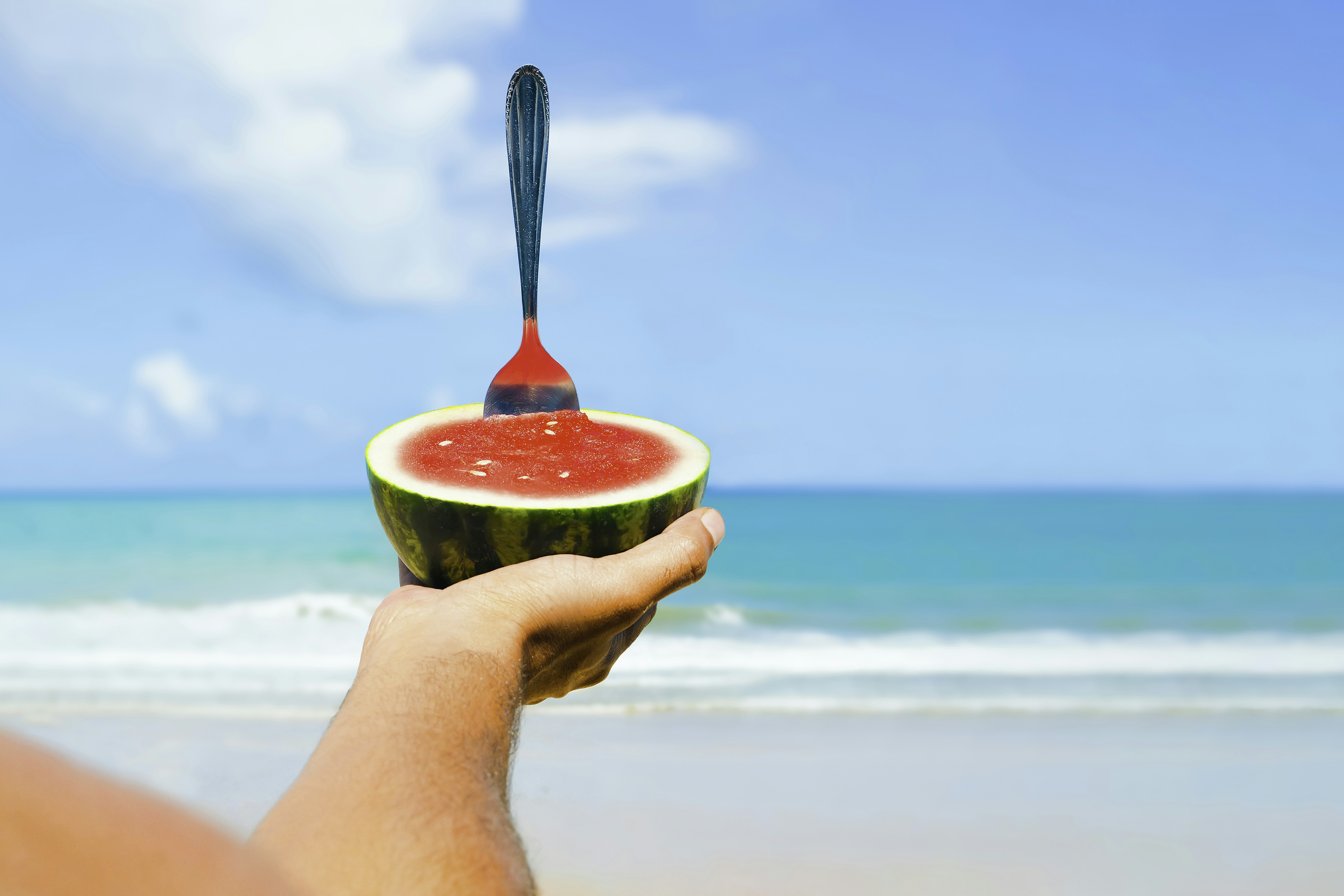 Bright hand holding a halved watermelon with a spoon against a turquoise sea and blue sky; the scene emphasizes fresh summer refreshment.
