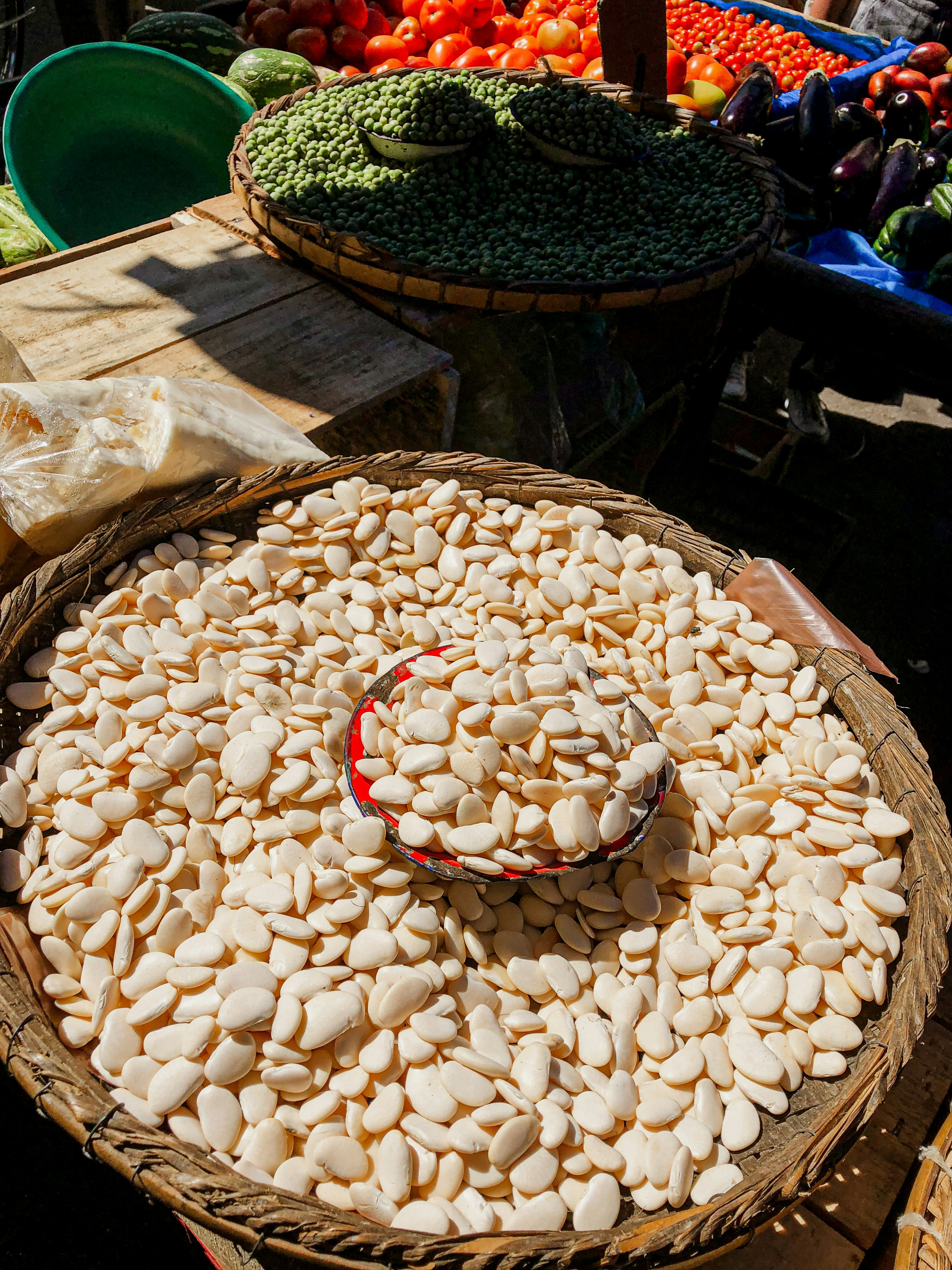 Sunlit market scene featuring a large woven basket heaped with pale seeds, a smaller ladle resting atop, with colorful produce in the background; a photography-ready still-life at a bustling outdoor stall. This is a photograph.