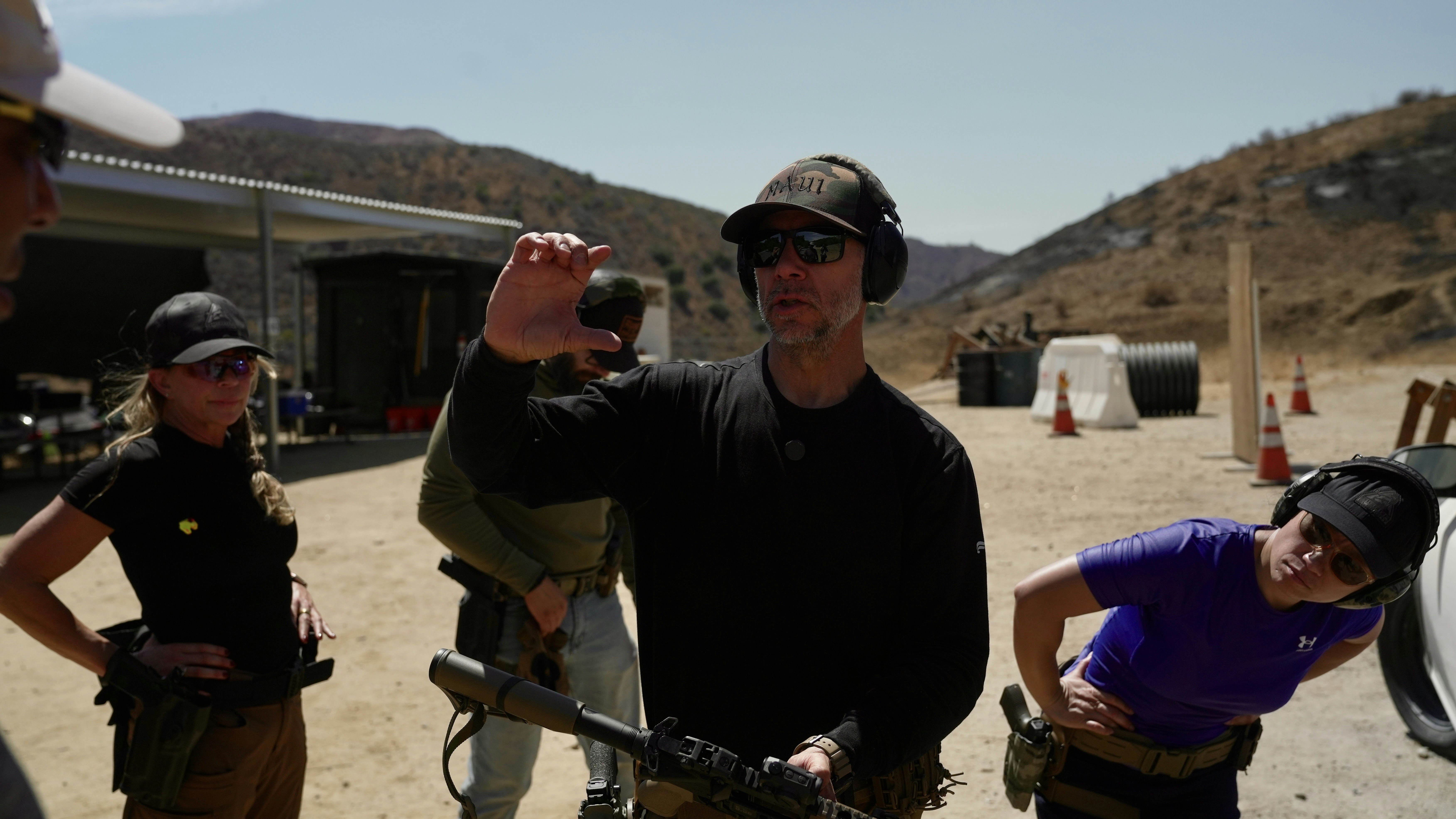 Instructor gestures with hand raised during a training drill amid a desert range, while teammates observe; the scene includes equipment and safety gear in a sunny outdoor setting.