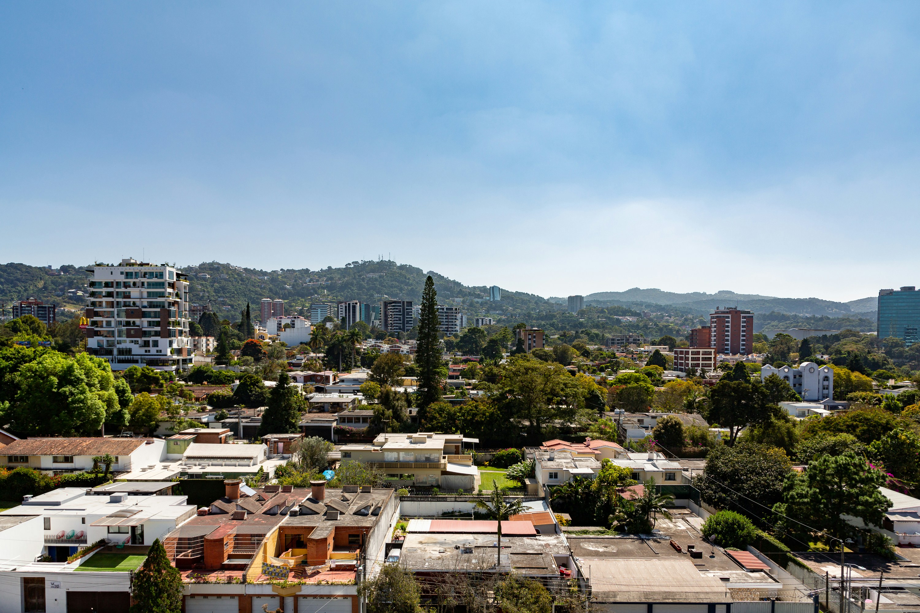 Sunlit cityscape with a grid of low-rise rooftops and scattered trees under a clear blue sky. This wide-angle view captures a tranquil urban edge with distant hills.