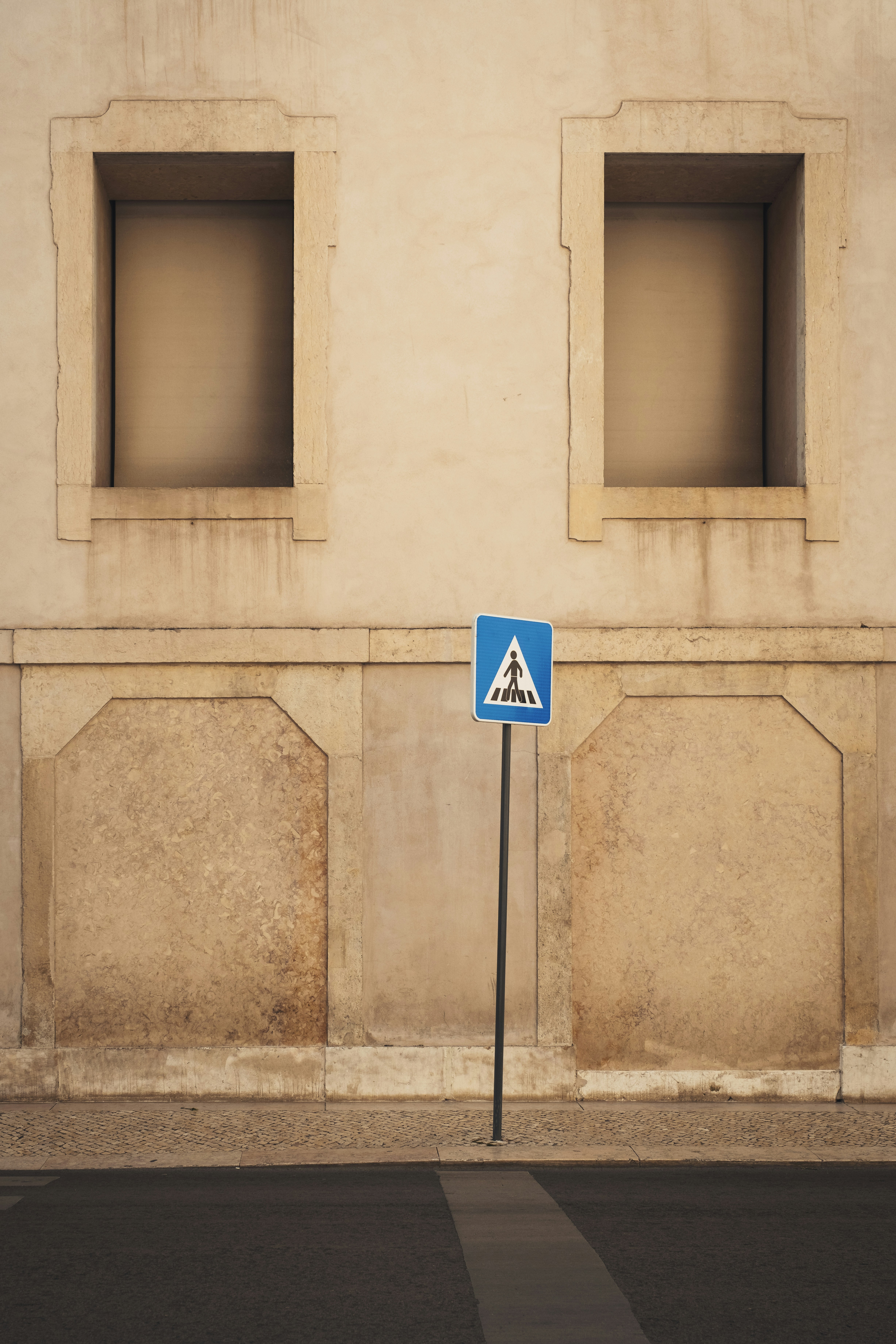 Beige wall with two square recessed windows and a lone pedestrian crossing sign centered along a weathered street.
