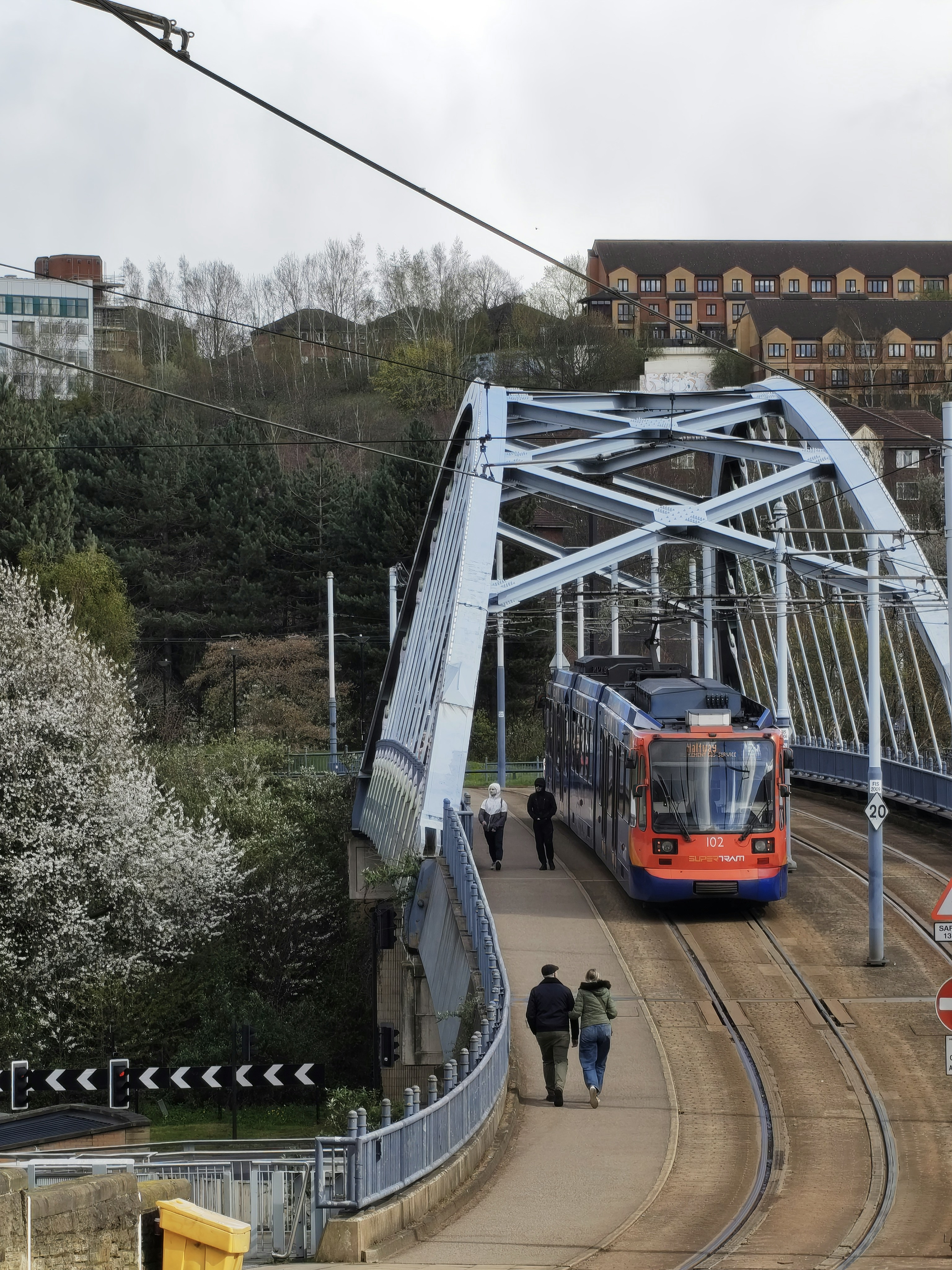 Red and blue tram crosses a curved blue-arch bridge with pedestrians on the path beside it; urban buildings and trees form the background.