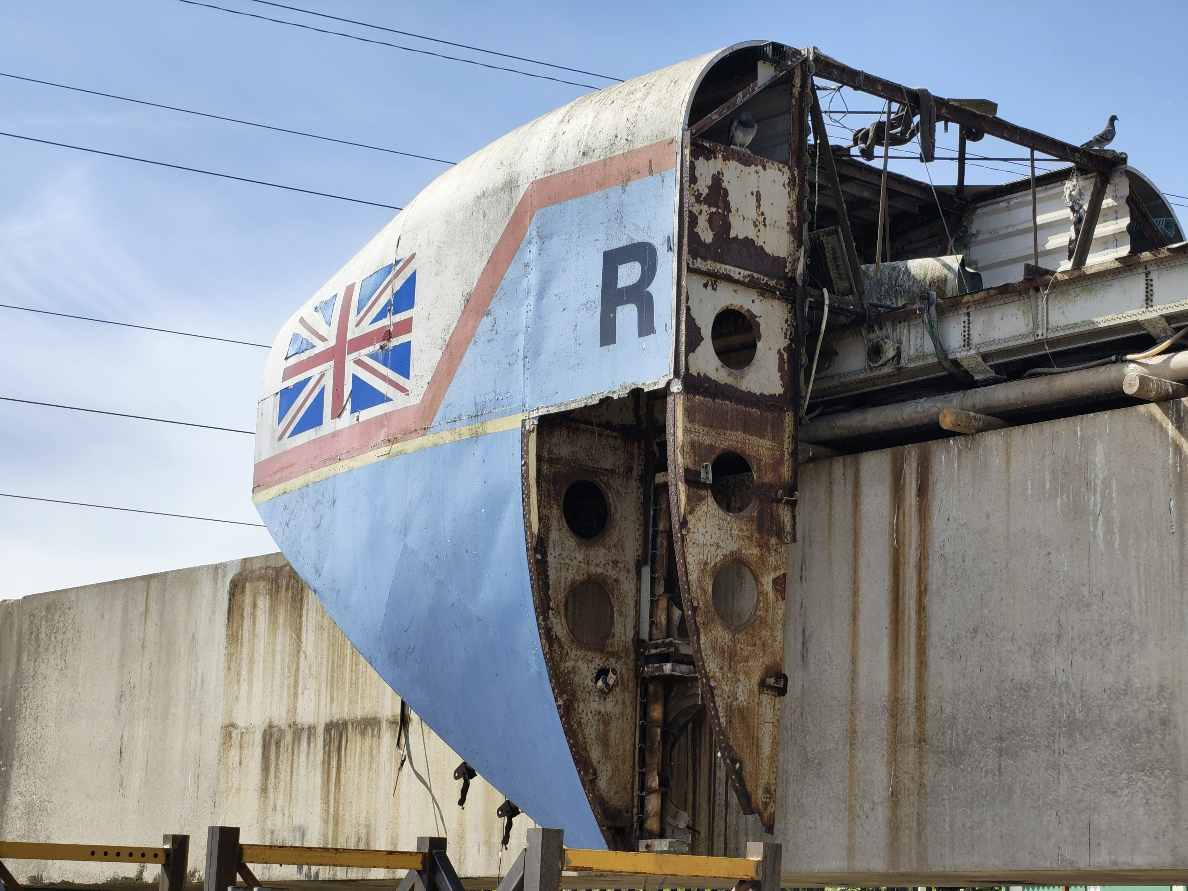 A weathered rocket nose sits on a rusty rail, bearing a faded Union Jack alongside a blue and white paint job; the scene highlights industrial decay and remnants of transport architecture. This is a photographic capture of a sculpture or derelict exhibit element at a port area.