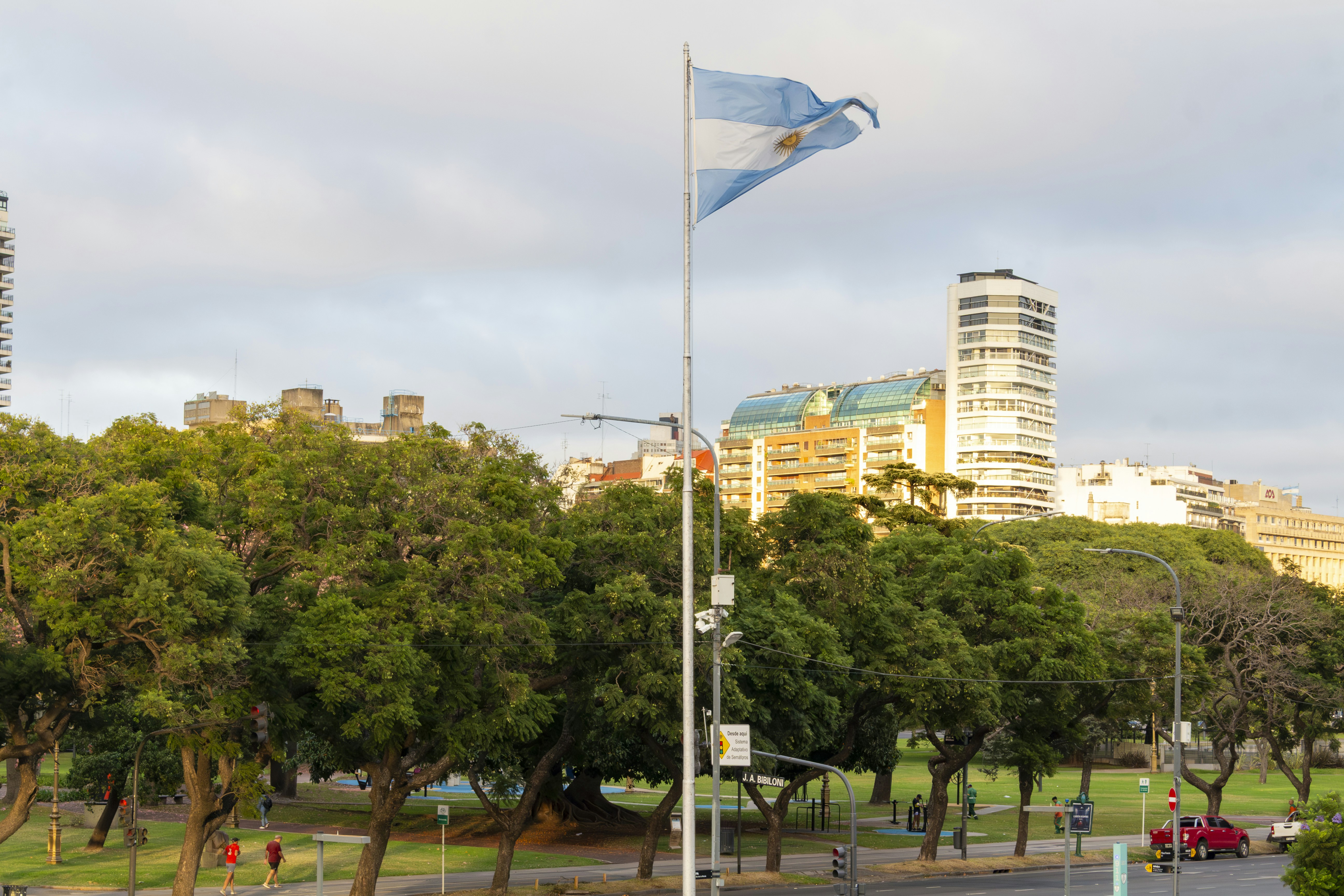 Flagpole and flag rise over a tree-lined park with urban buildings in the background, capturing a calm city scene.