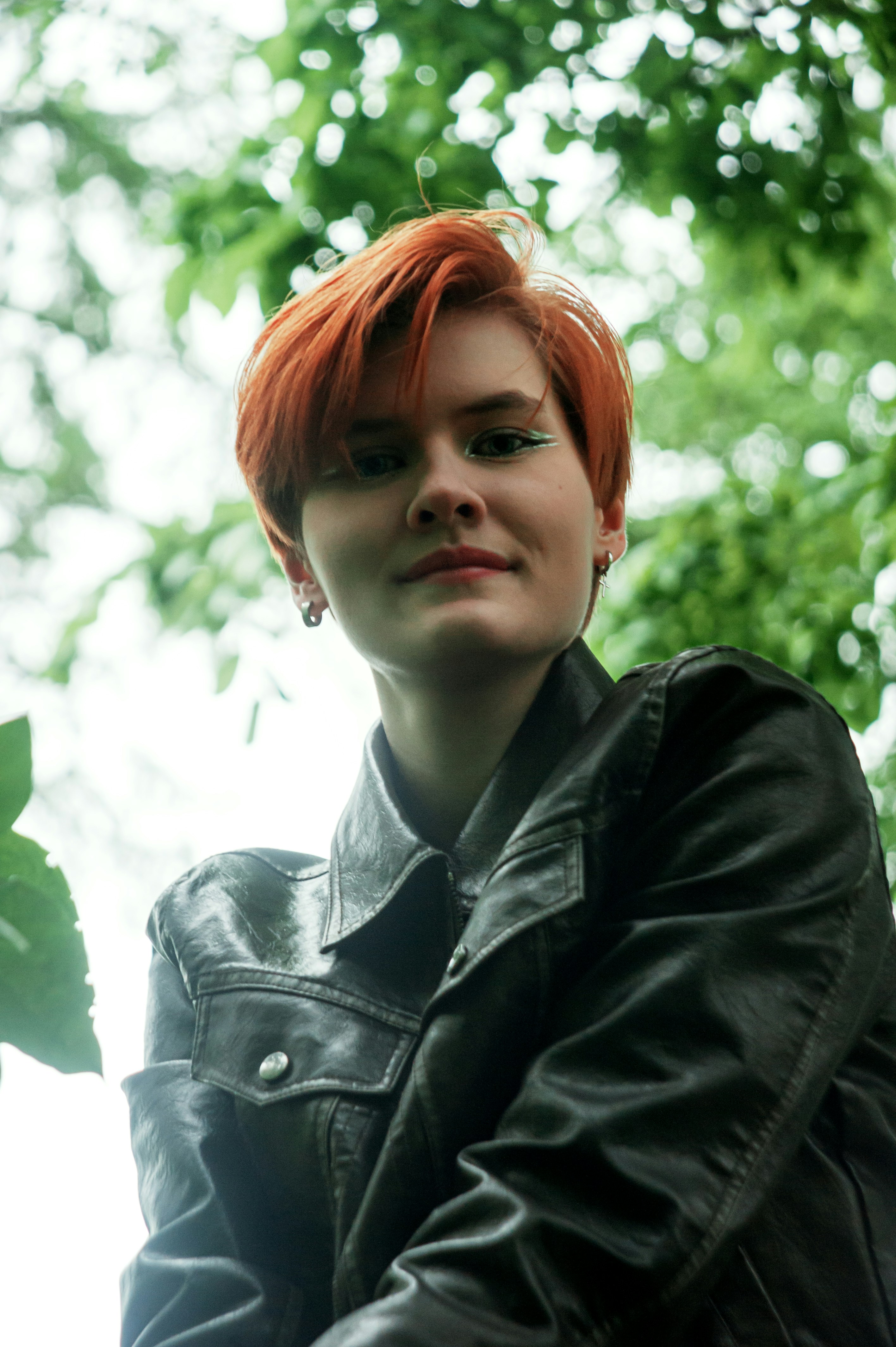 Portrait of a person with short red hair wearing a black leather jacket against a lush green background; the image emphasizes their confident expression and natural light.
