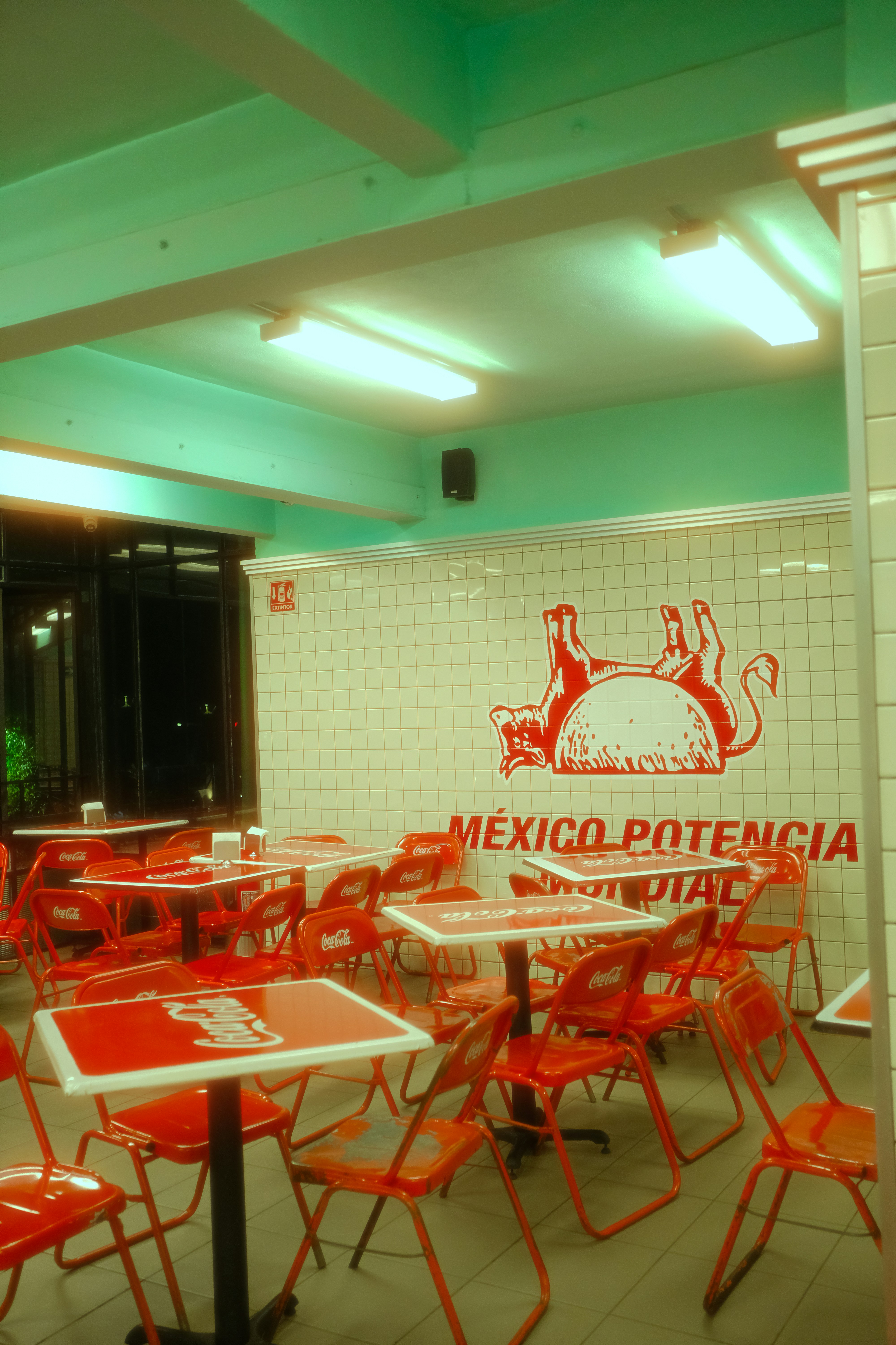 Bright red Coca-Cola chairs and tables line a tiled wall with a bold red mural and Mexican text in a casual dining area.