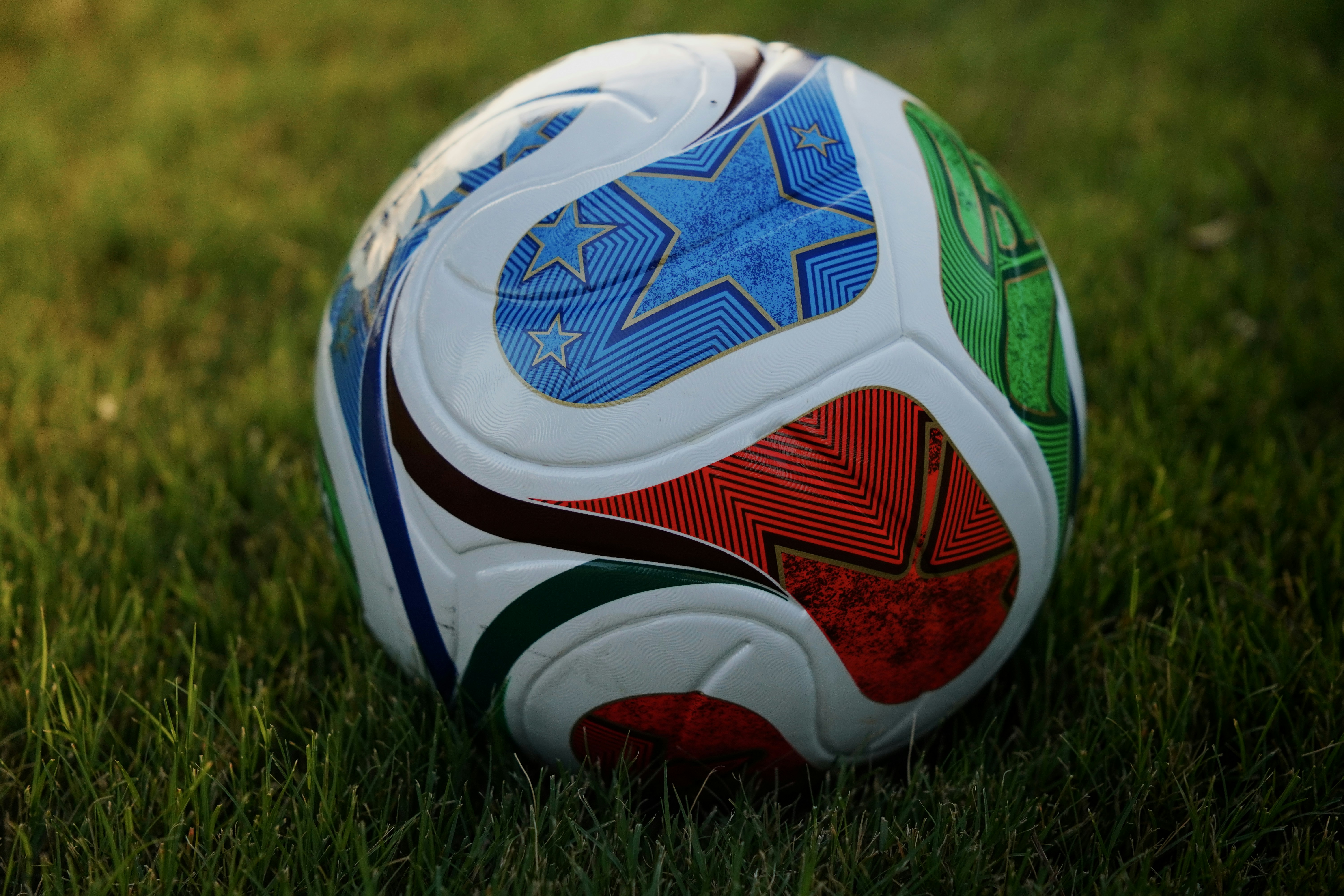 Bright patterned soccer ball sits close to the grass, with vivid blue, red, green, and white panels catching soft outdoor light. The club-style ball appears ready for play on a damp field.