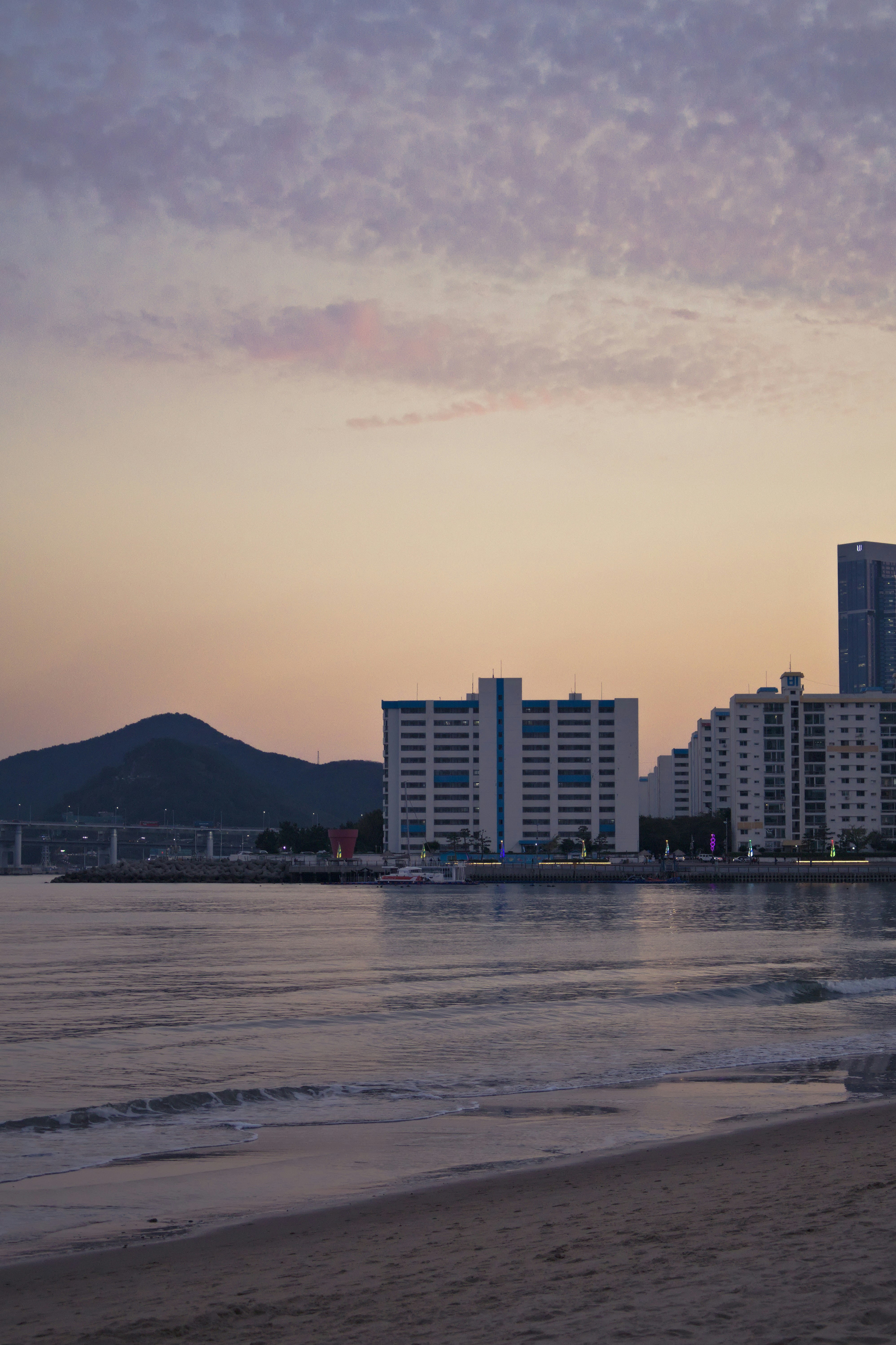 Soft golden sunset washes the city coastline while calm water leads to benched high-rise buildings and a distant hill, capturing urban silhouettes against a pastel sky. The scene emphasizes the tranquil beach context with a distant urban horizon.