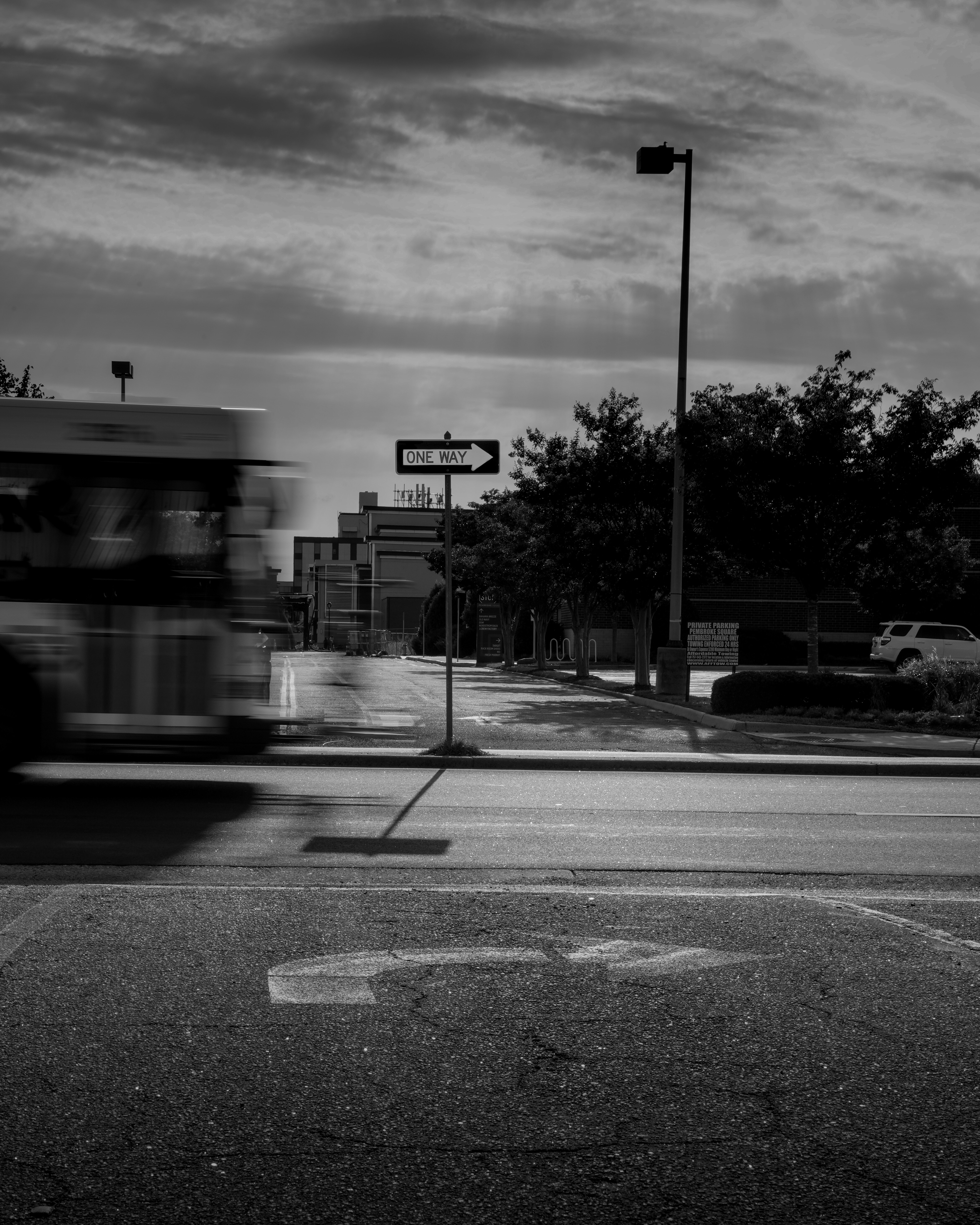 Street scene with a blurred vehicle crossing in the foreground and a clear sidewalk and buildings in the background; a crosswalk arrow marking and a one-way sign anchor the composition, conveying motion and urban rhythm.