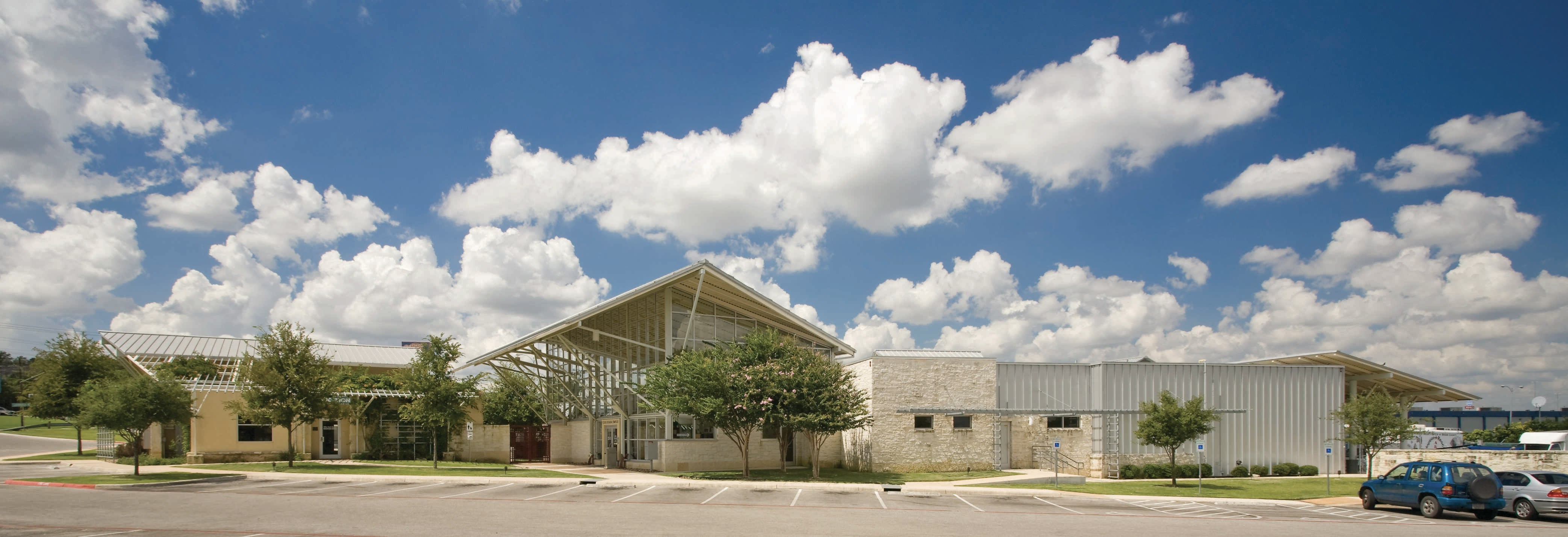 Wide exterior of a single-story civic/retail building with a peaked entry and glass canopy, set under a bright blue sky; a storefront campus with a parking area and a blue vehicle visible on the right. This is a architectural/commercial exterior scene.