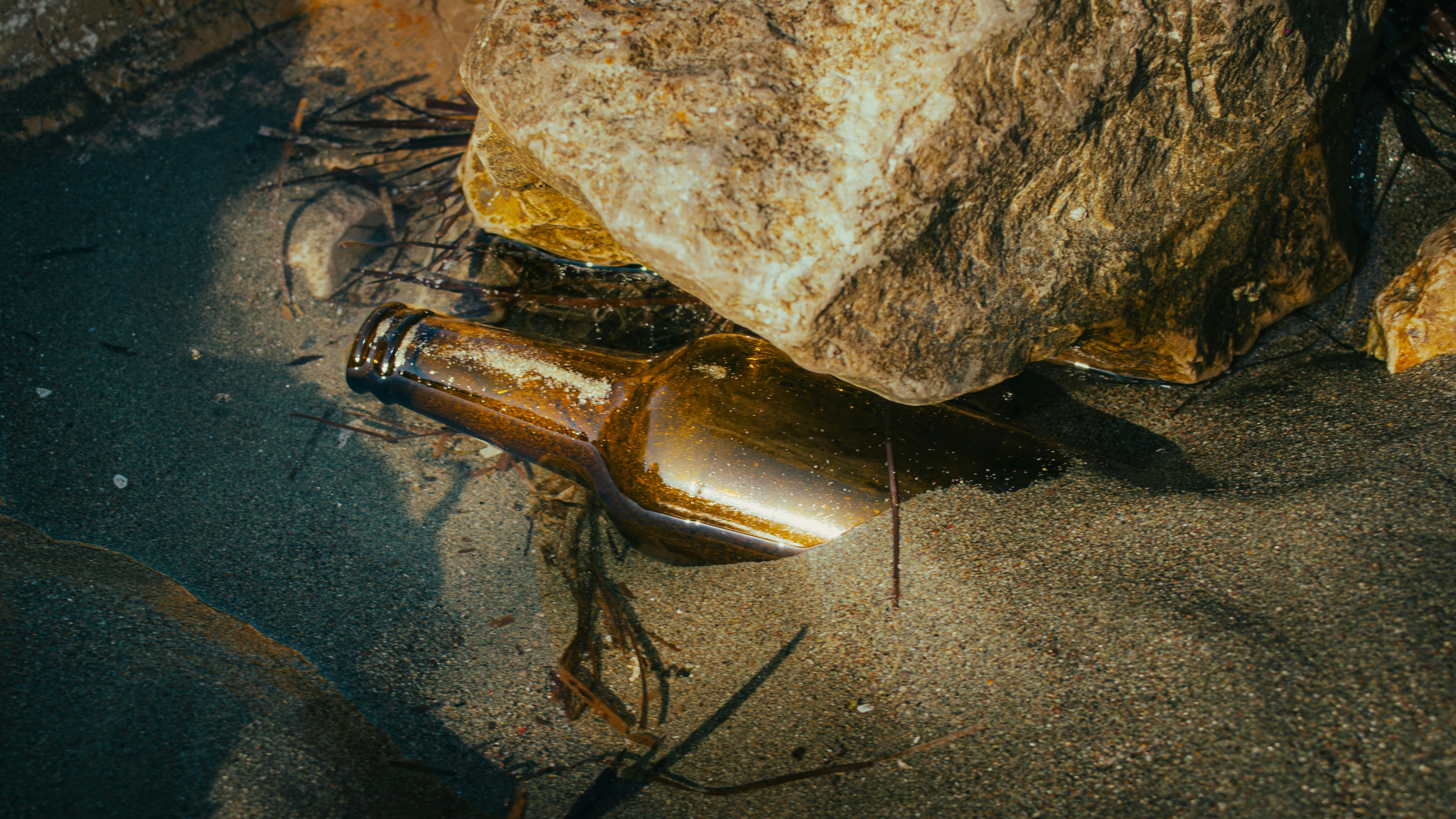 Rusty bottle partly buried under rock along a dark, sandy shore; minimal waves and seaweed hint at a forgotten detritus scene. (Not a logo, illustration, painting, or cartoon.)