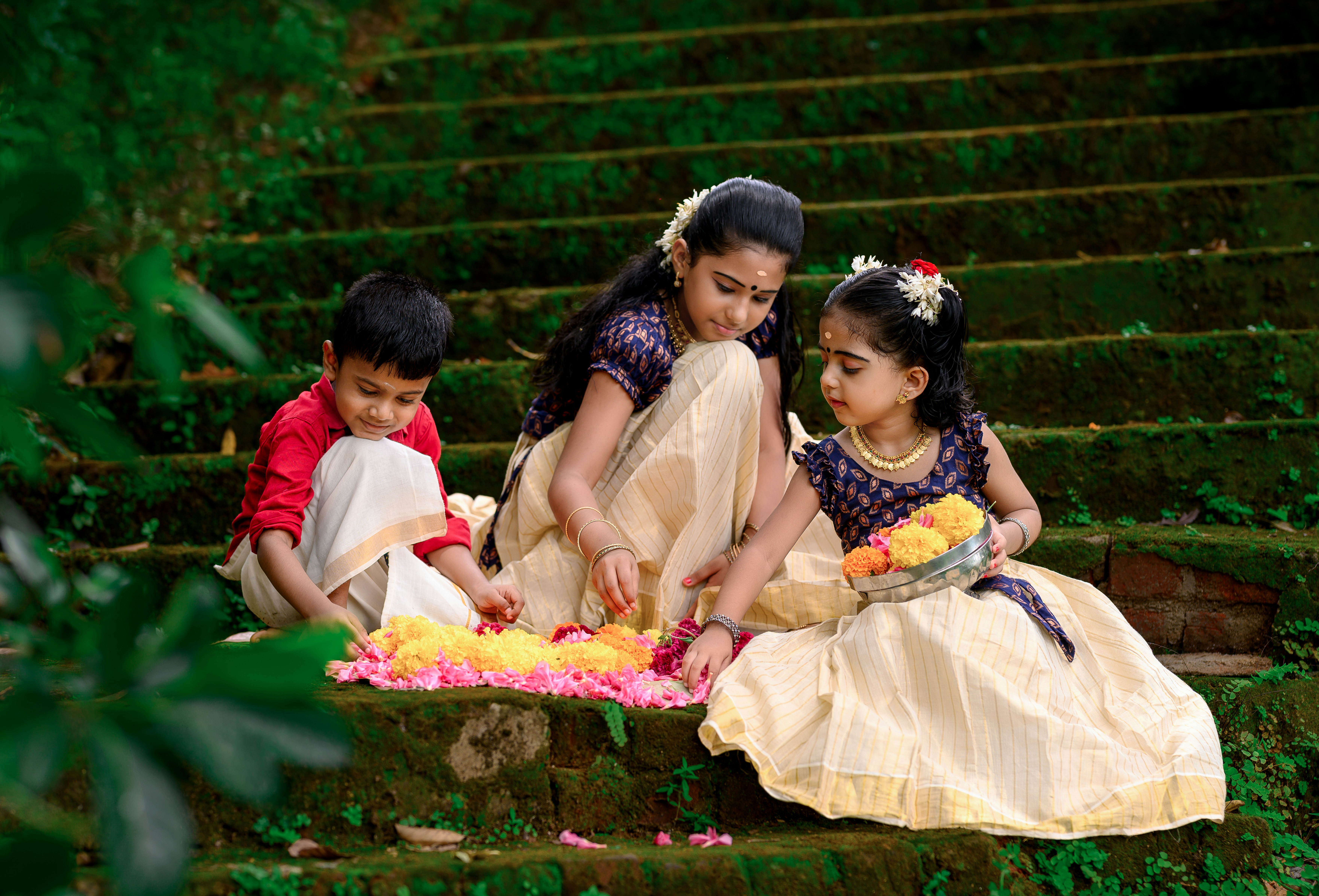 Three children in colorful traditional outfits arrange flower petals on stone steps; a candid familial moment with natural greenery in the background.