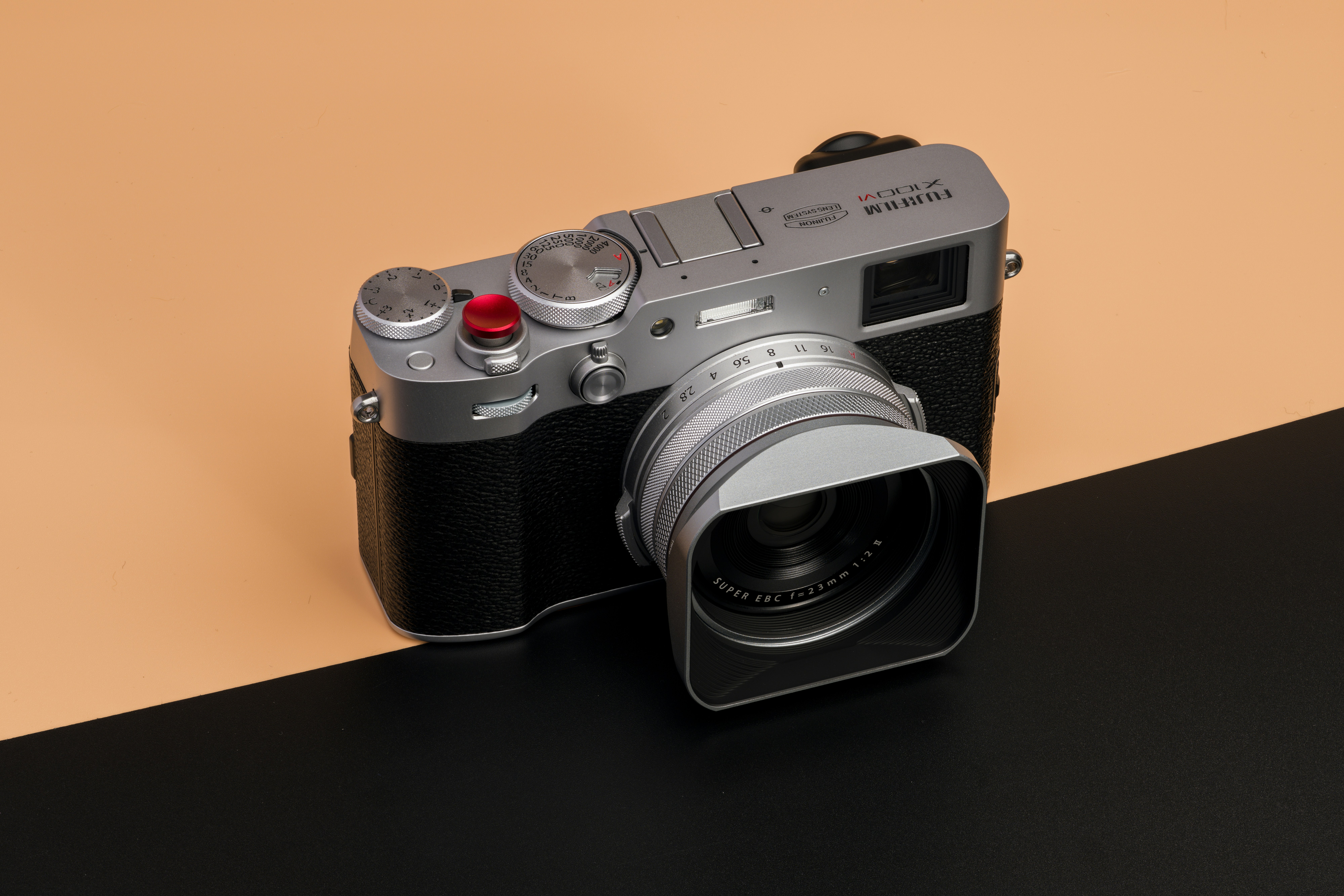 Close-up of a vintage-style Nikon camera with a silver body and black textured grip resting on a two-toned backdrop; a compact lens with a rectangular hood dominates the foreground.