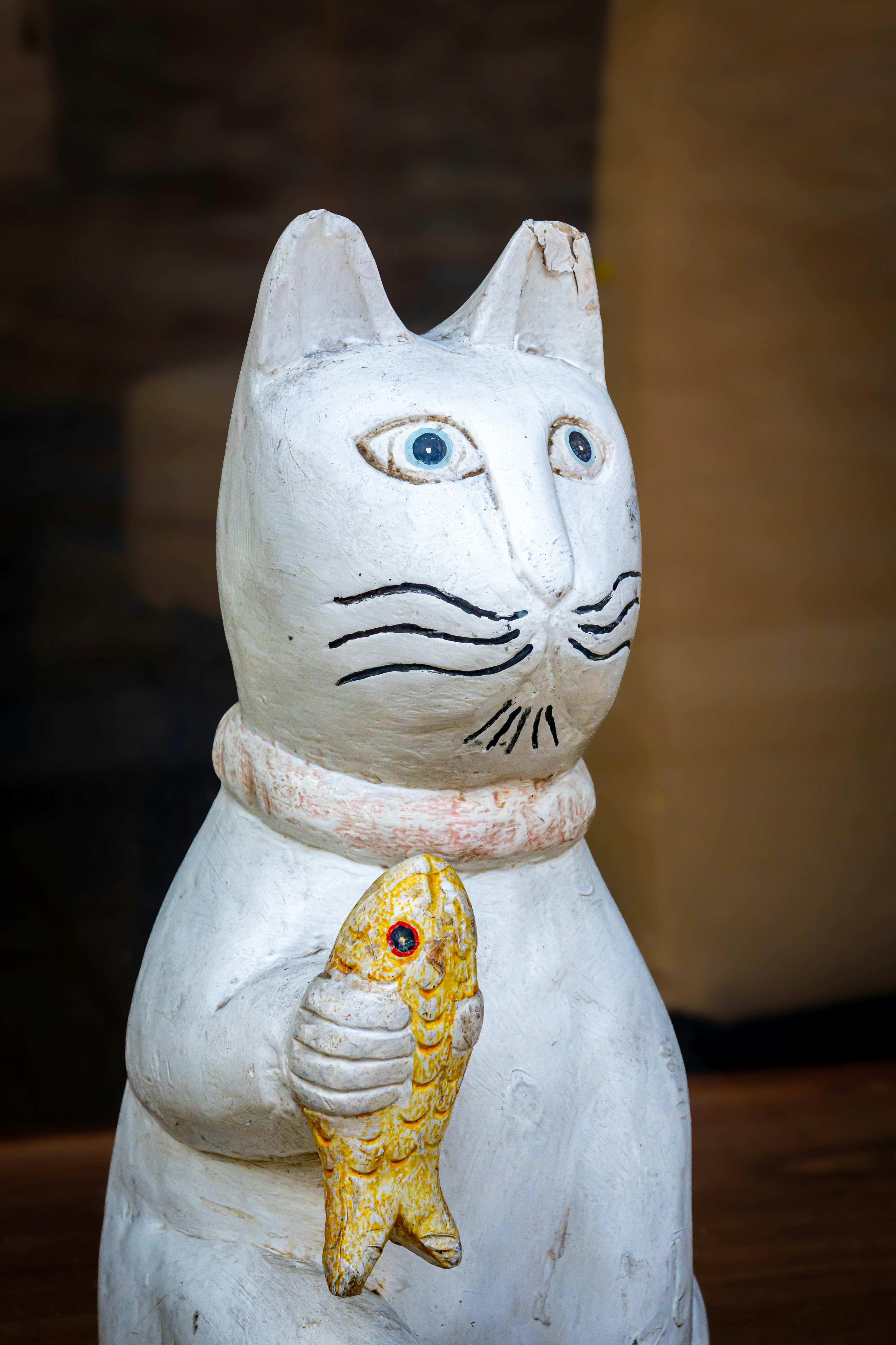 White ceramic cat with a yellow fish in its paw stands in warm shadowed room