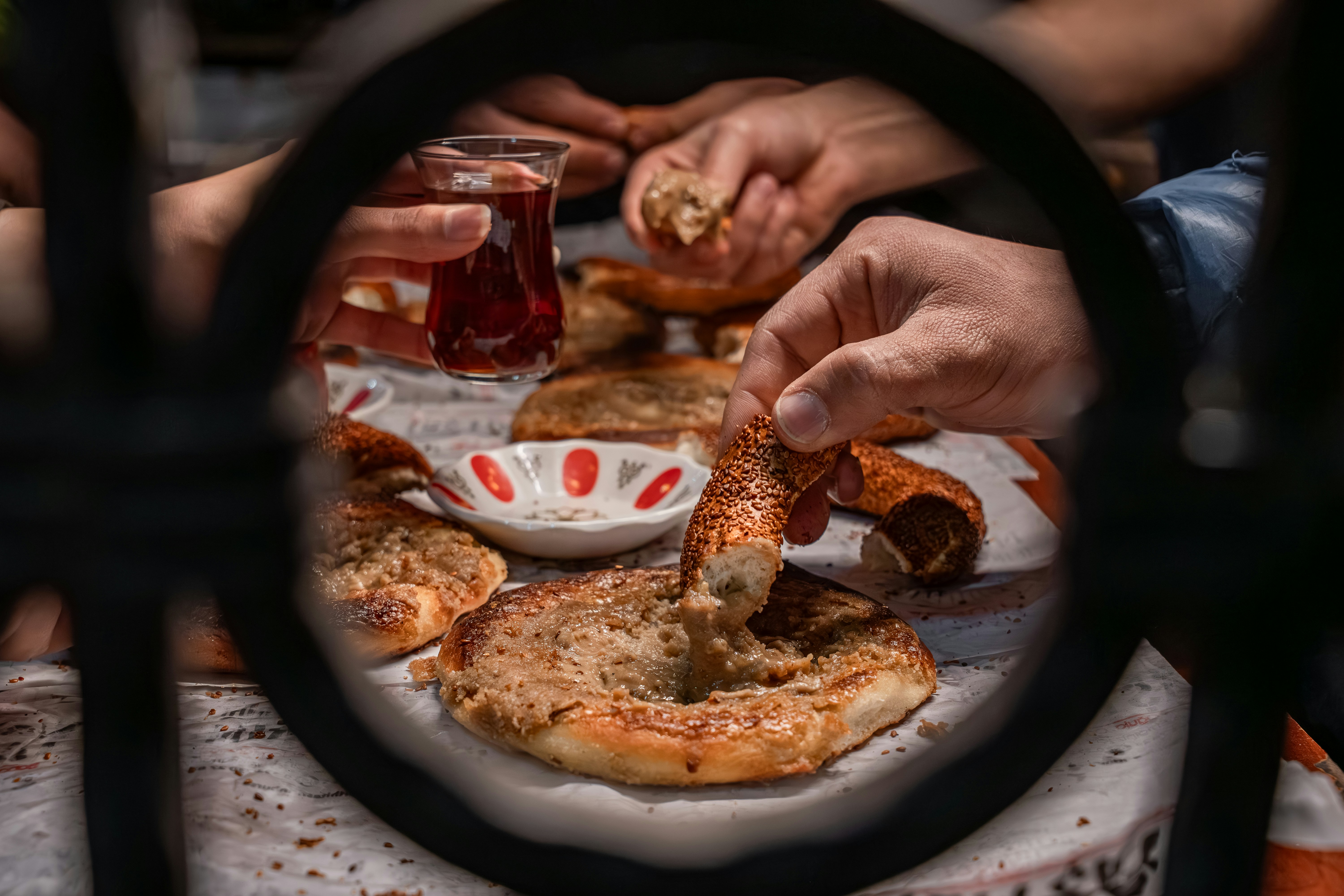 Close-up of hands dipping sesame bread into a plate of sauce and tearing bread, framed by outstretched arms and circular foreground elements from a kitchen setting.