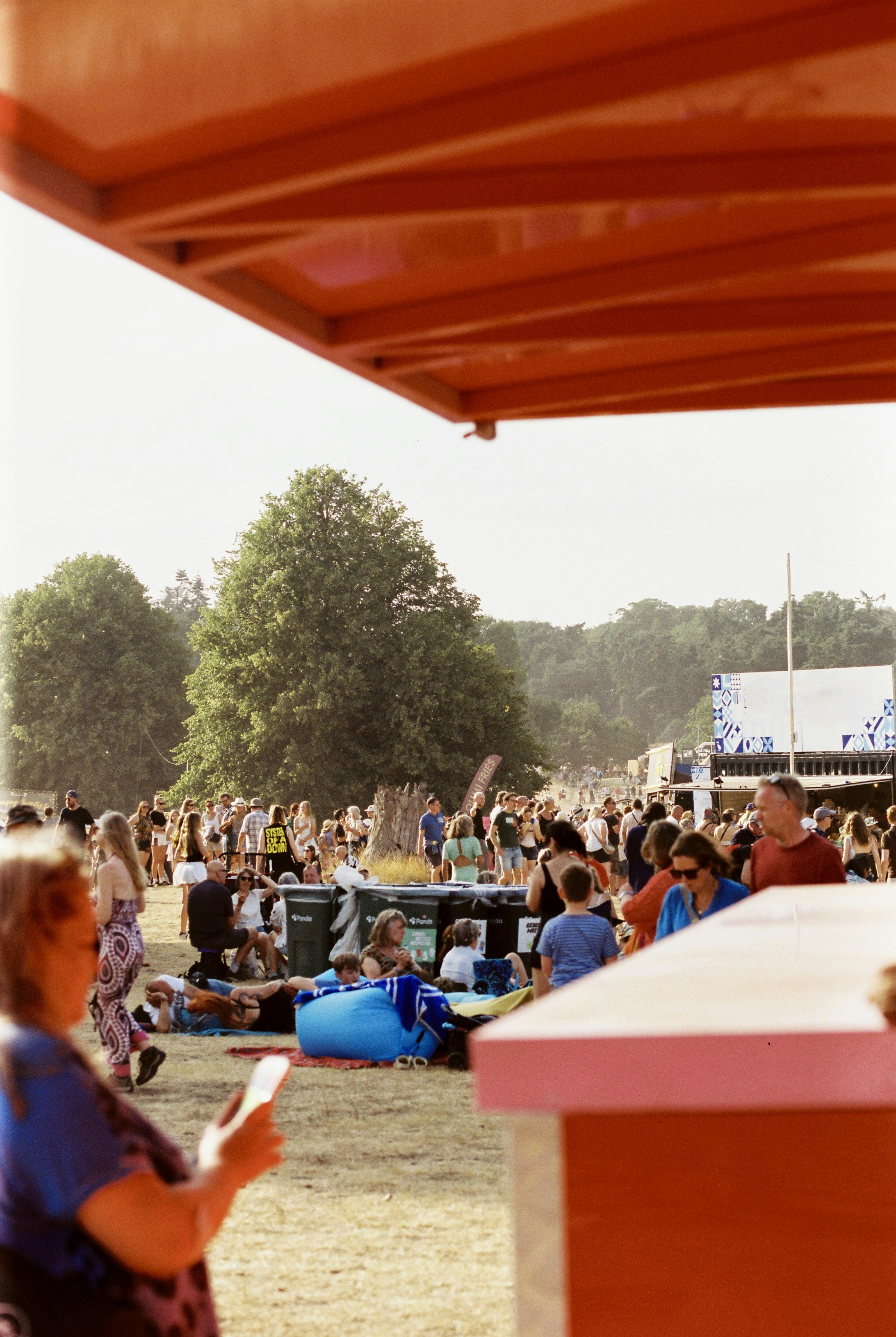 Crowd gathered at an outdoor festival with a stage seen in the distance; people relaxing on the grass, trees in the background, and a wooden structure in the foreground.