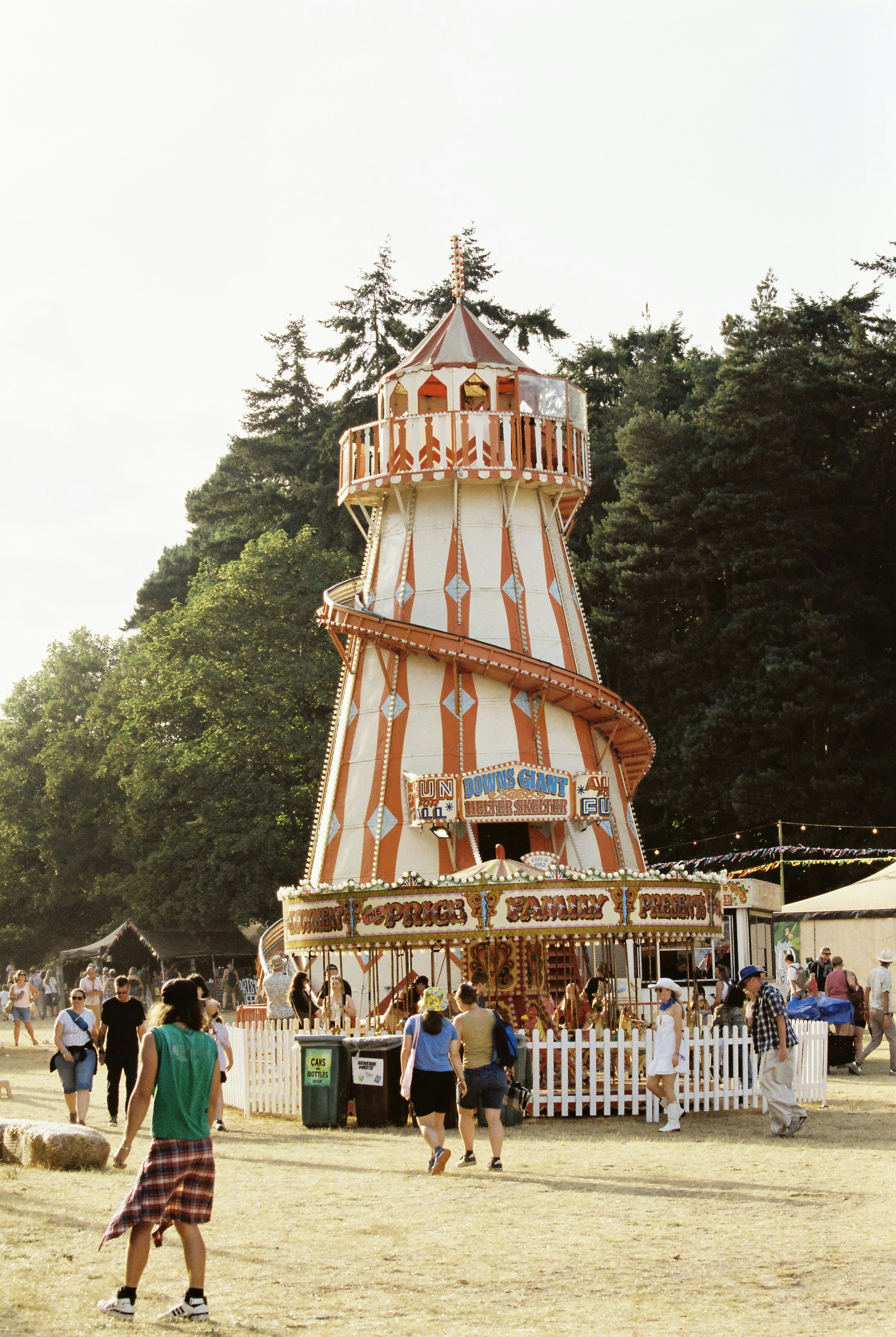 Bright striped carousel tower rising above a lively fairground with visitors and trees in the background. A nostalgic, outdoor scene capturing summer activity.