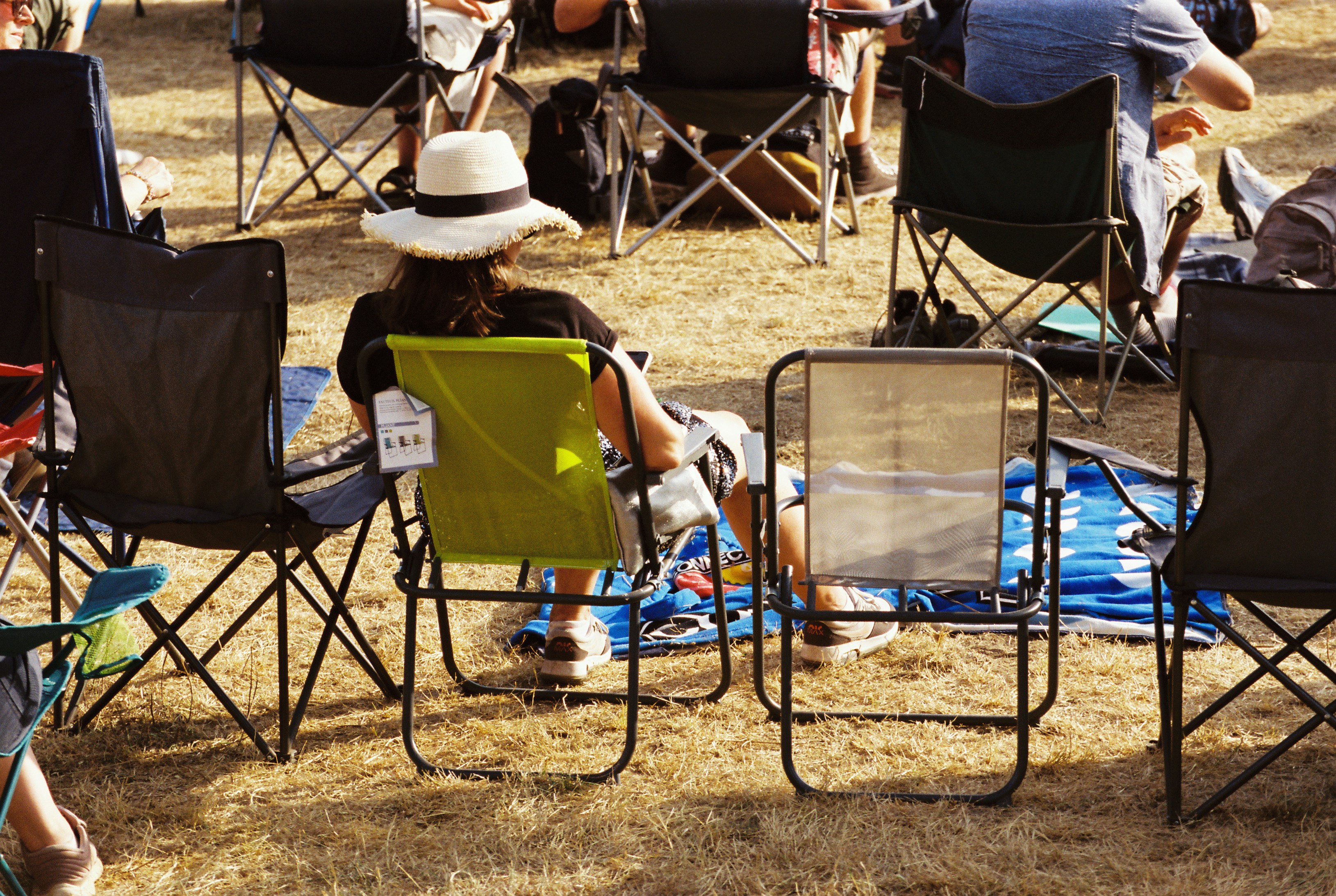 Sunlit outdoor event with attendees seated in folding chairs on dry grass, captured from behind a row of chairs toward the crowd.