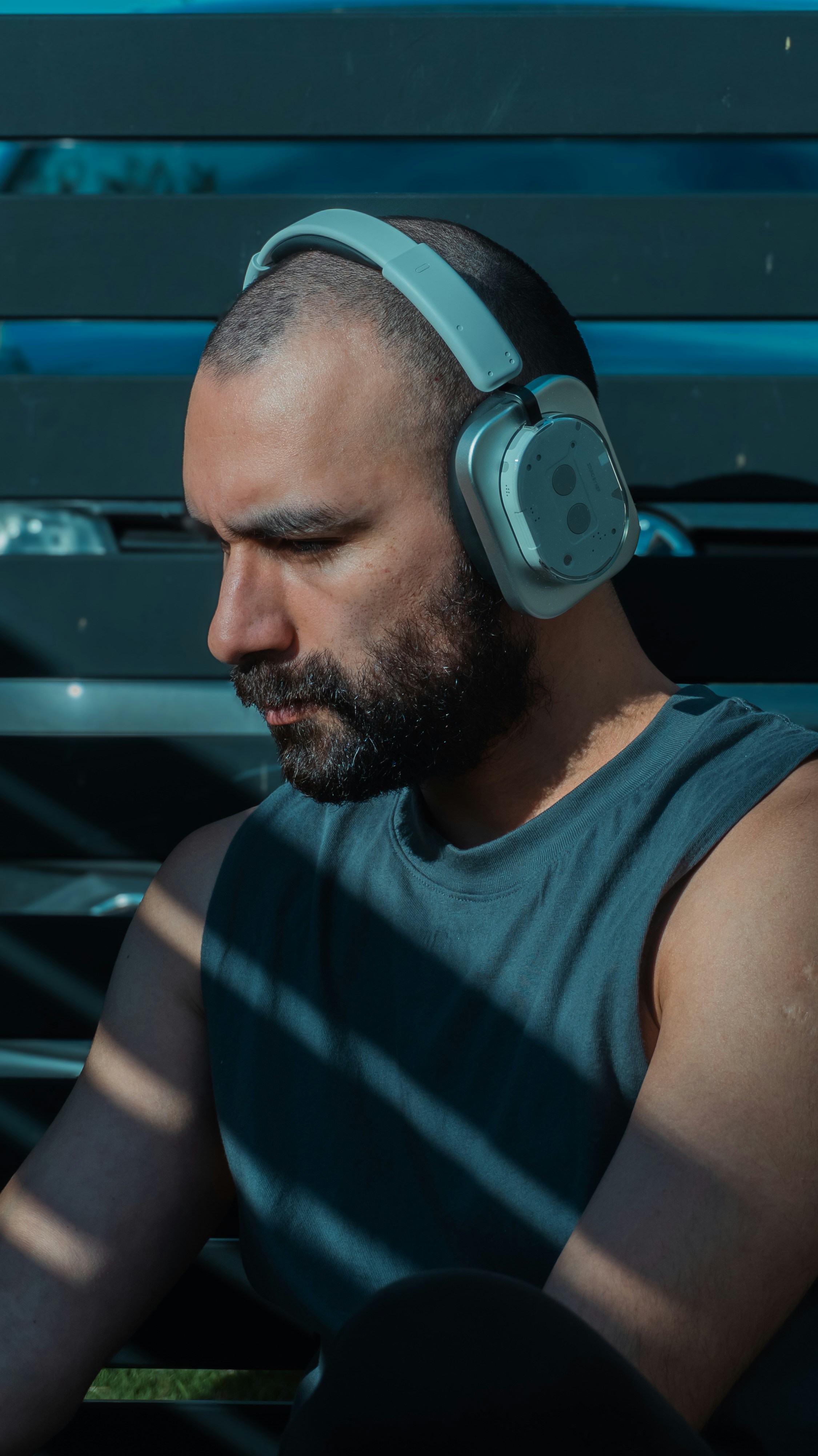 Senior man wearing teal over-ear headphones sits against a dark blue backdrop; dramatic shadows highlight facial features and beard. Photograph captures a focused, introspective moment with a portrait-like vibe. (c) logo-free, color-graded portrait.