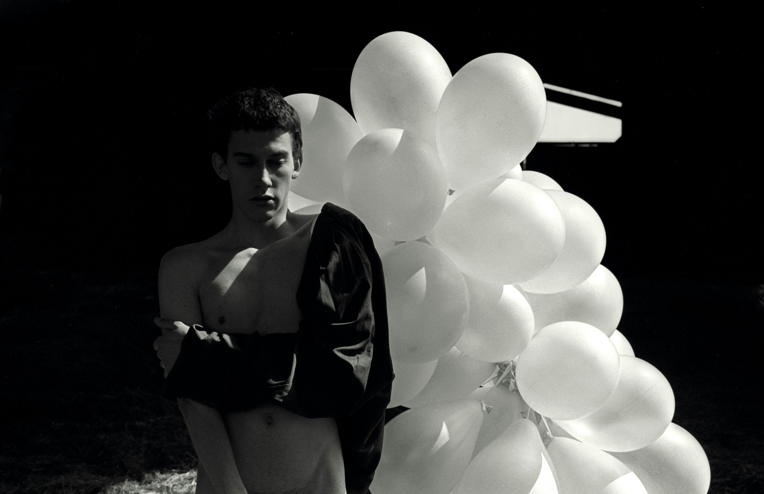 Young man stands shirtless in shadow next to a cluster of white balloons, creating a stark, monochrome silhouette.