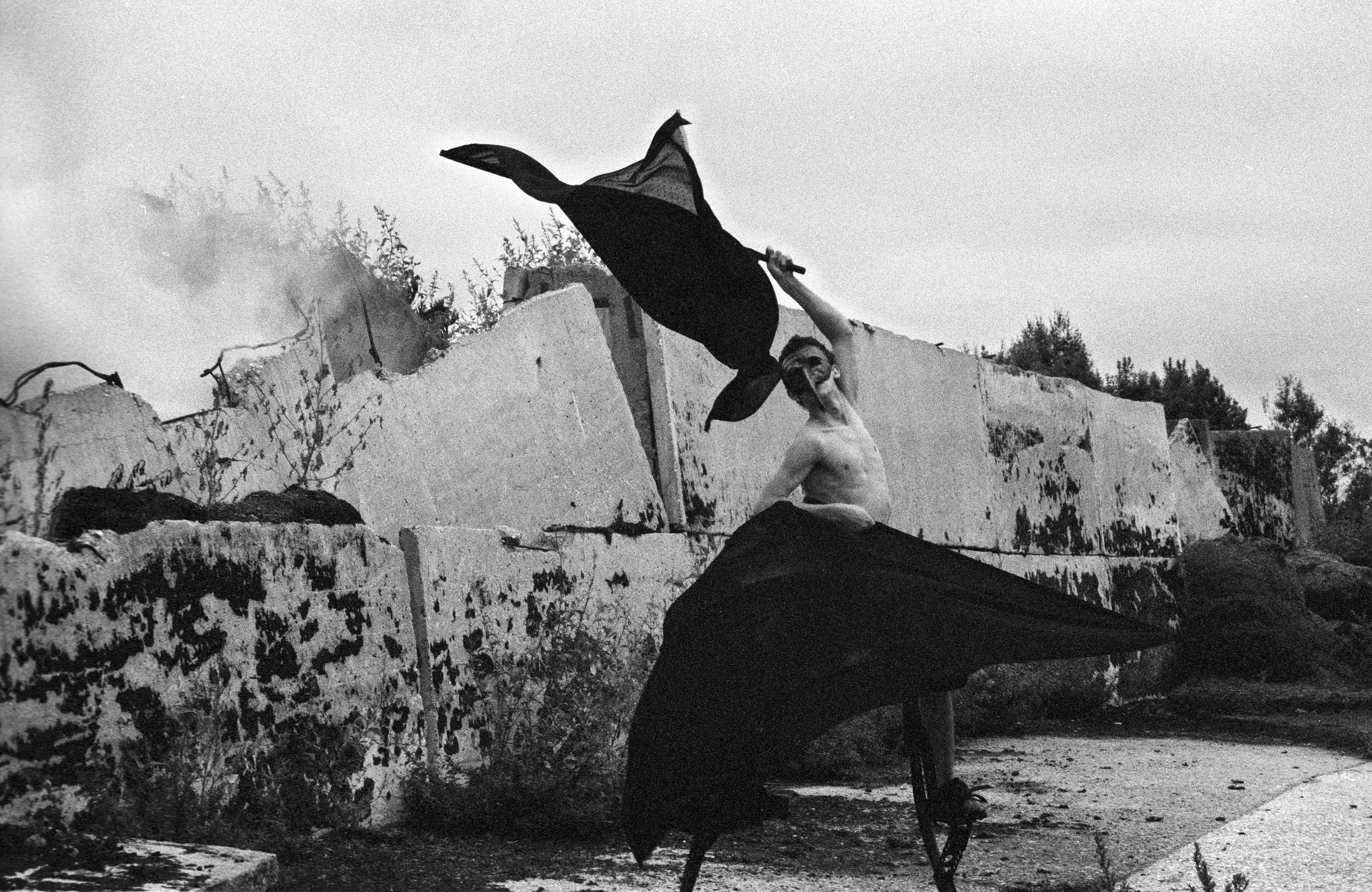 A figure in motion stands by a textured concrete seawall with a flowing dark fabric, captured in a grainy monochrome. The scene prioritizes the dynamic pose and architectural backdrop.