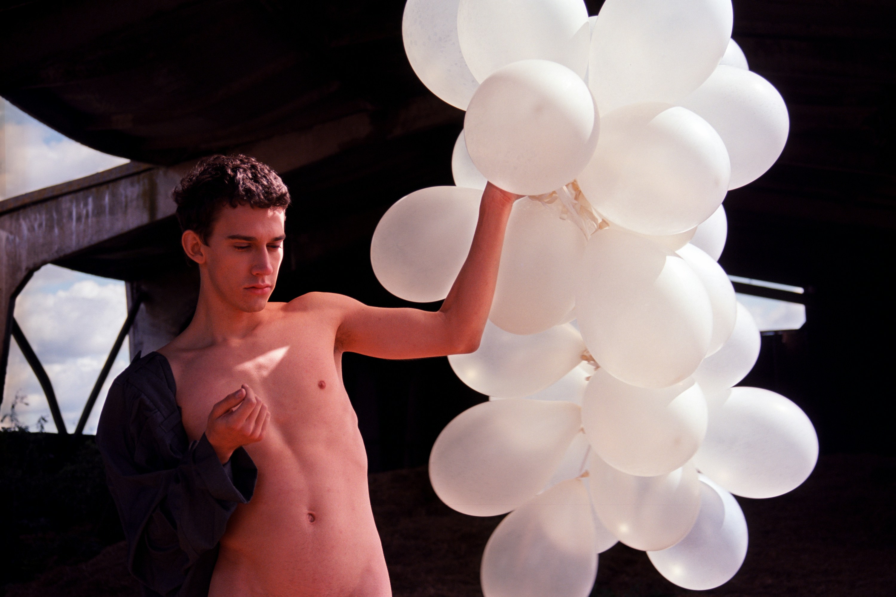 A shirtless young man stands under a cluster of white balloons, gazing downward as he holds the balloons with one arm in a relaxed, intimate pose. A dark industrial background contrasts with the soft, airy balloons.