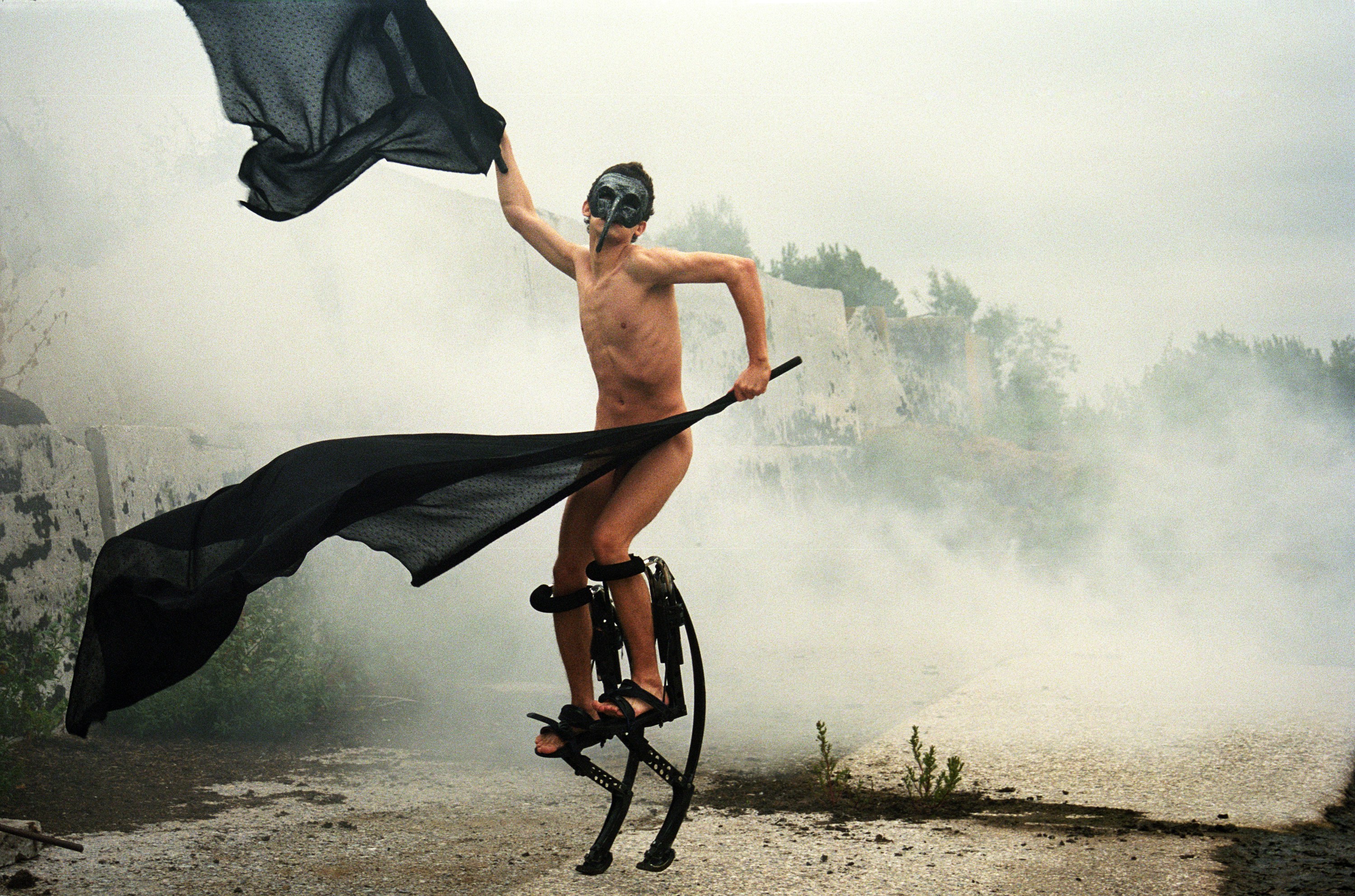 A shirtless performer on a skeletal bike surfs through a smoky, desolate landscape while black fabric billows overhead, creating a dramatic contrast and motion.