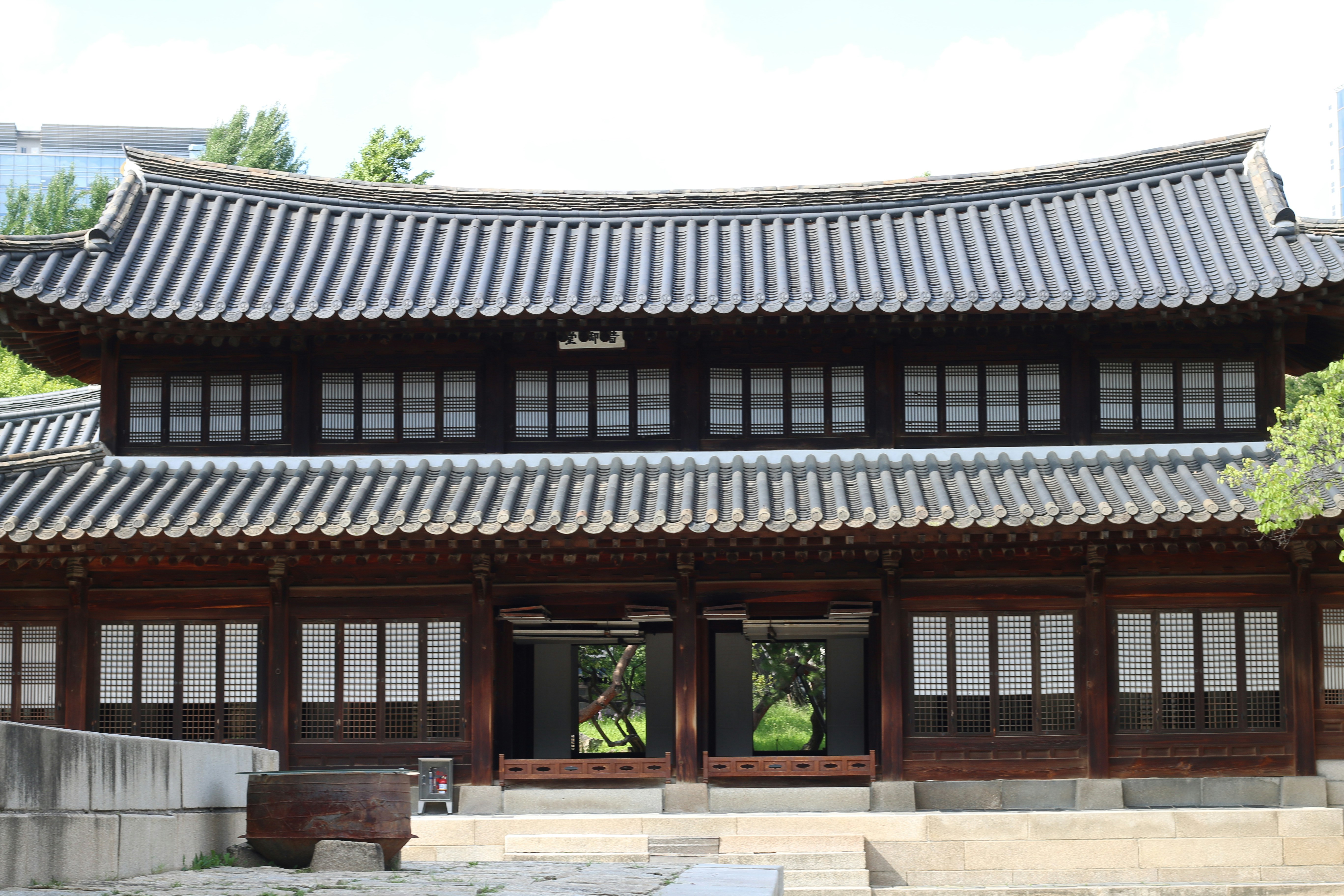 Traditional wooden hanok with a tiled roof and open central doorway reflecting a green courtyard; a calm architectural scene emphasizing symmetry and historical detail.