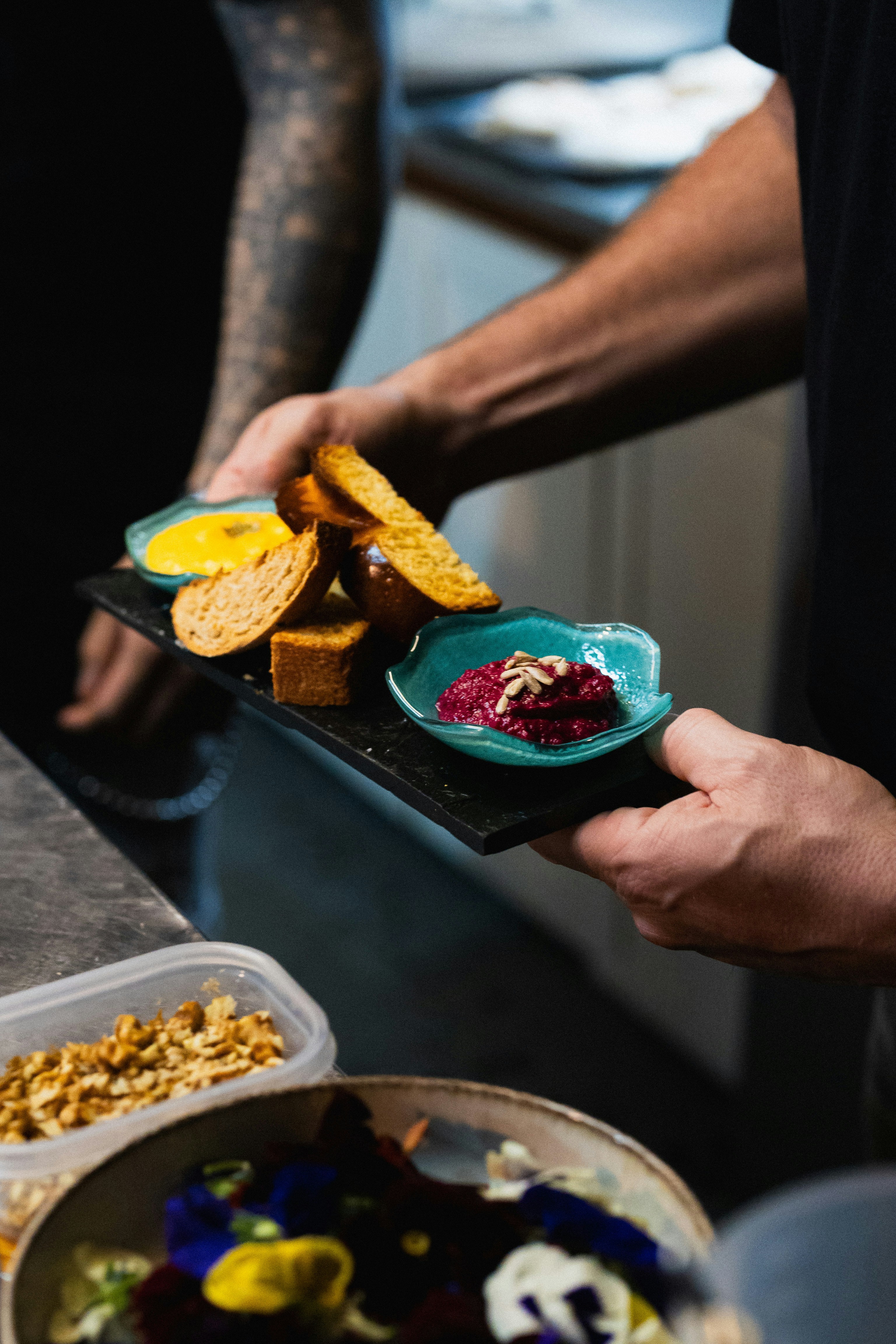 Close-up of a server presenting a small tray with toasted bread and colorful spreads; a kitchen scene hinting at a plated appetizer. (Logo/branding not visible.)