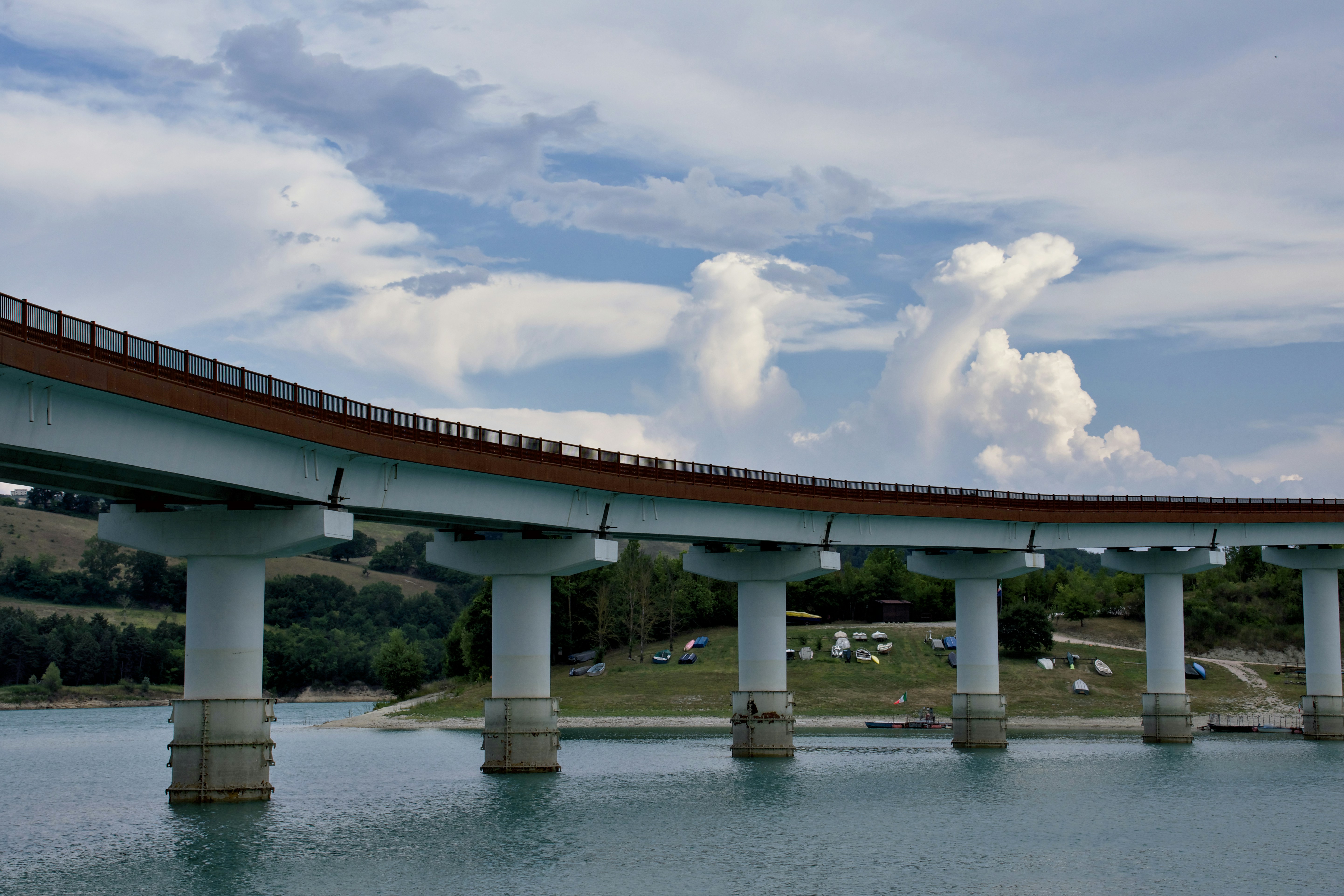 Wide-angle view of a curved bridge spanning a calm river, supported by tall pillars and a grassy riverbank visible in the distance.