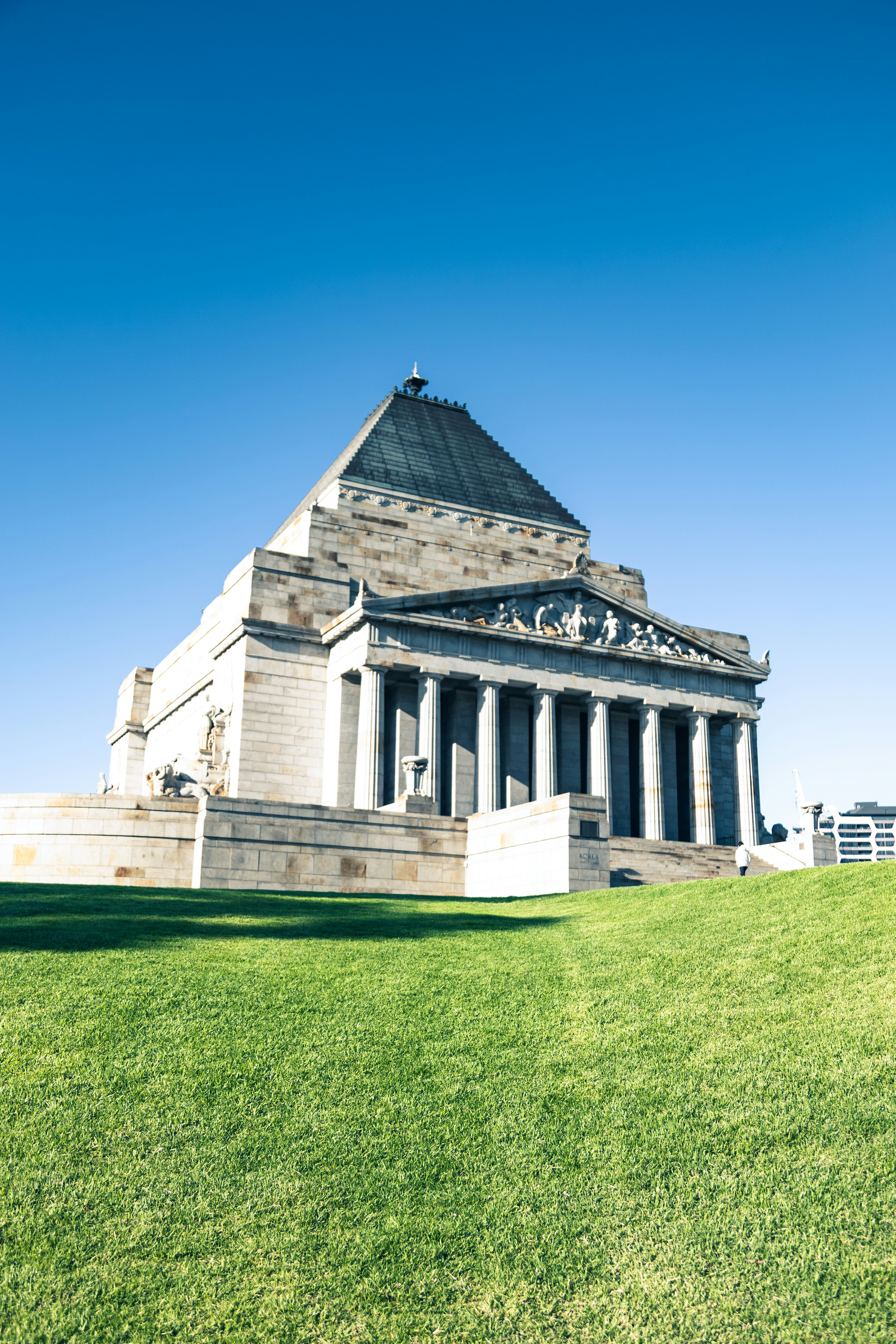 A stone monument with a pyramidal roof sits atop a stepped podium, flanked by a colonnade, as a lush green lawn stretches in the foreground under a bright blue sky.