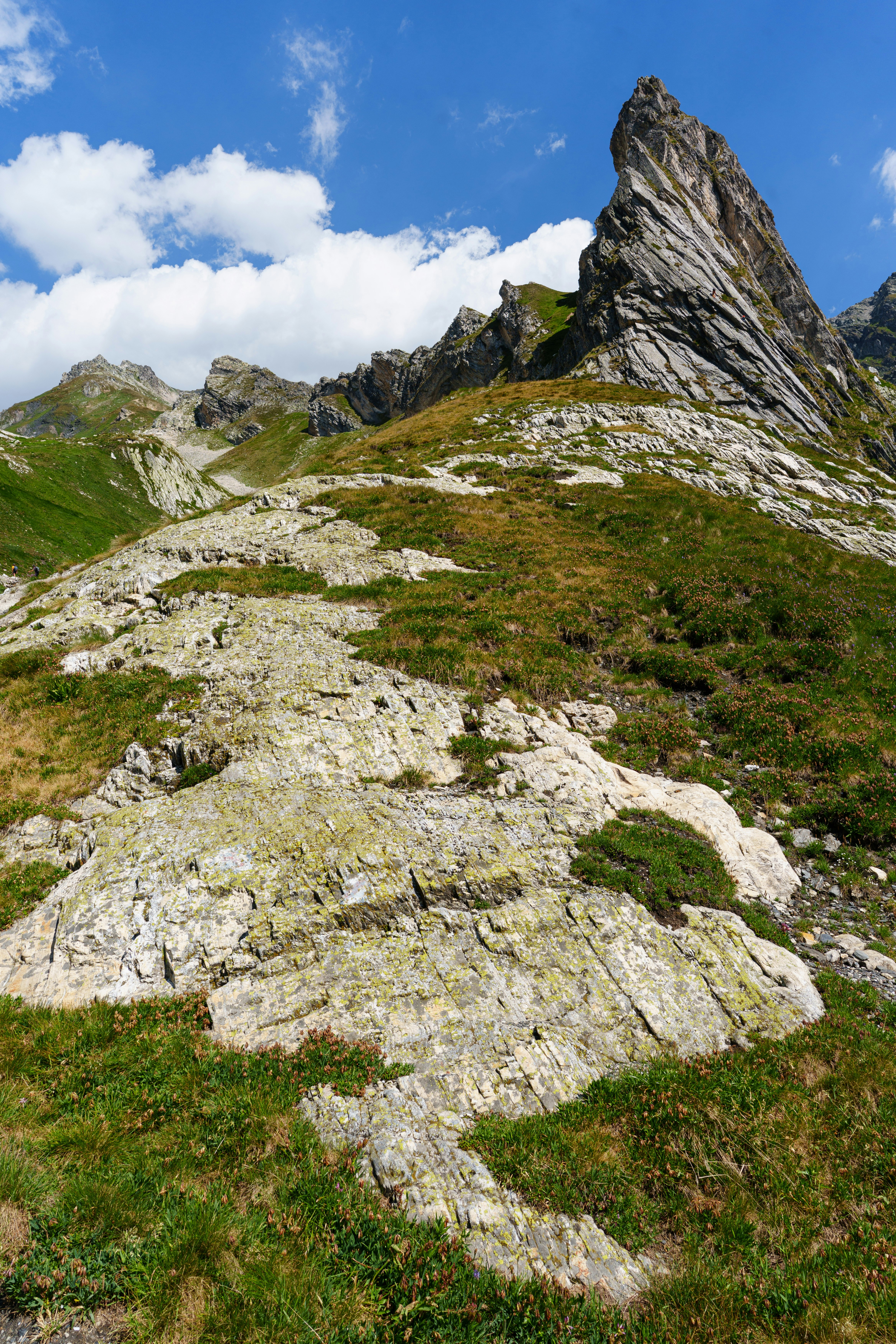 Sloping rocky terrain and a jagged pinnacle reach toward a bright blue sky with scattered clouds, showcasing a alpine landscape with patches of green moss and hardy shrubs.