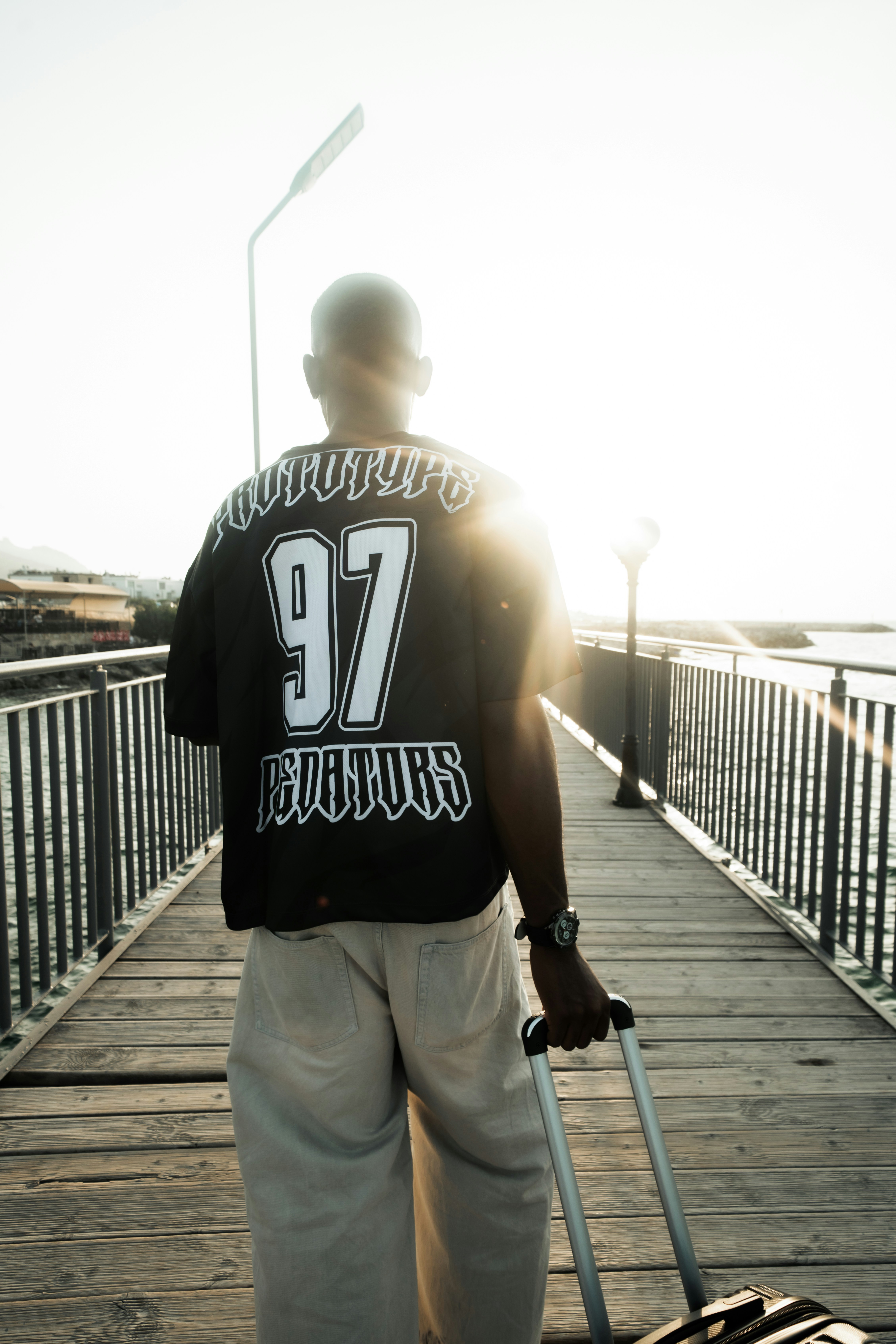 Back of a man with a large '97' jersey standing on a wooden pier, pulling a rolling suitcase toward a bright horizon. The scene emphasizes silhouette and urban-adventure mood. (Logo/graphic on jacket visible.)