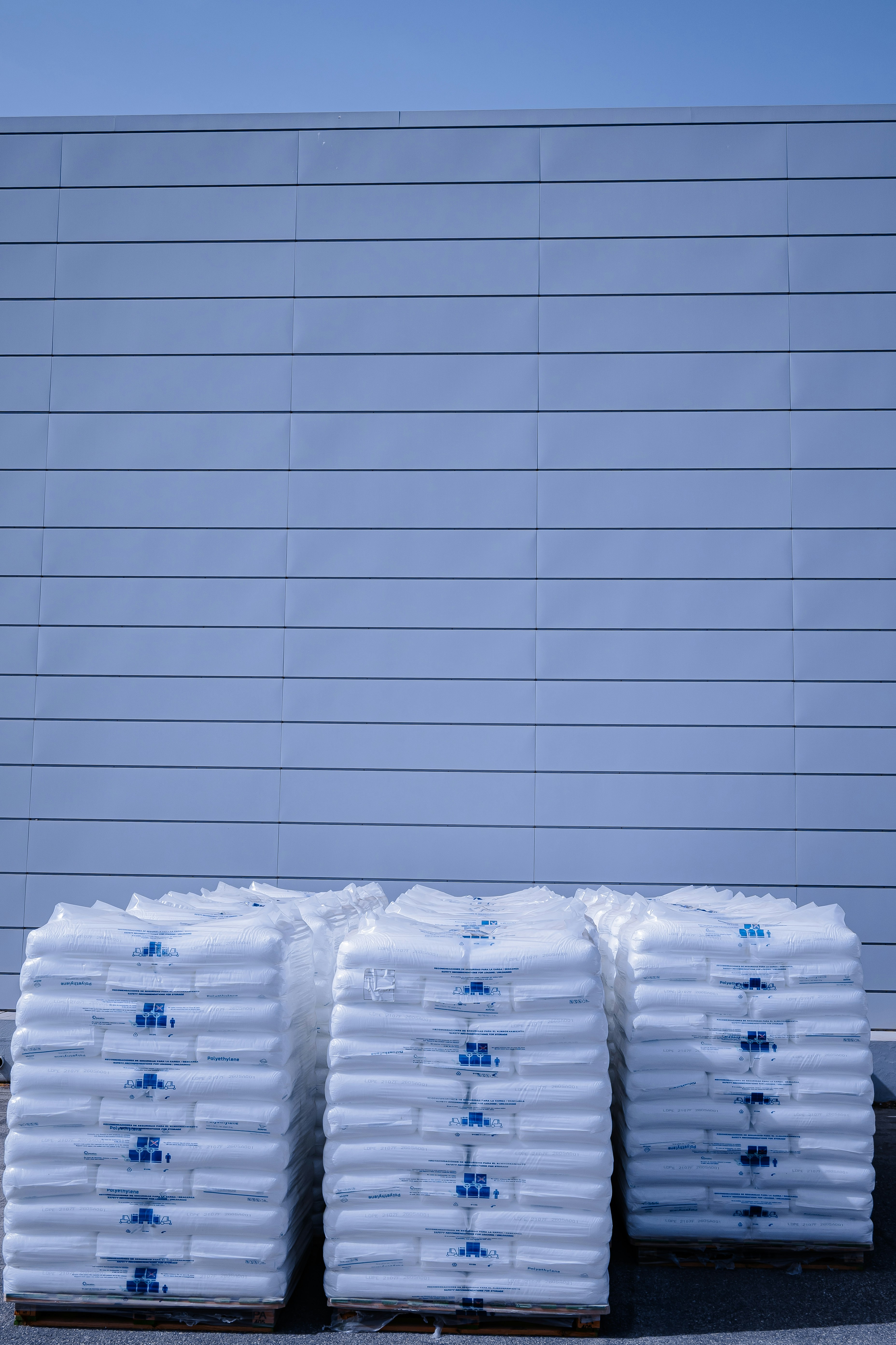 Stacked white bags sit in neat columns against a pale blue industrial wall, with a clear sky above and a flat, empty ground plane.