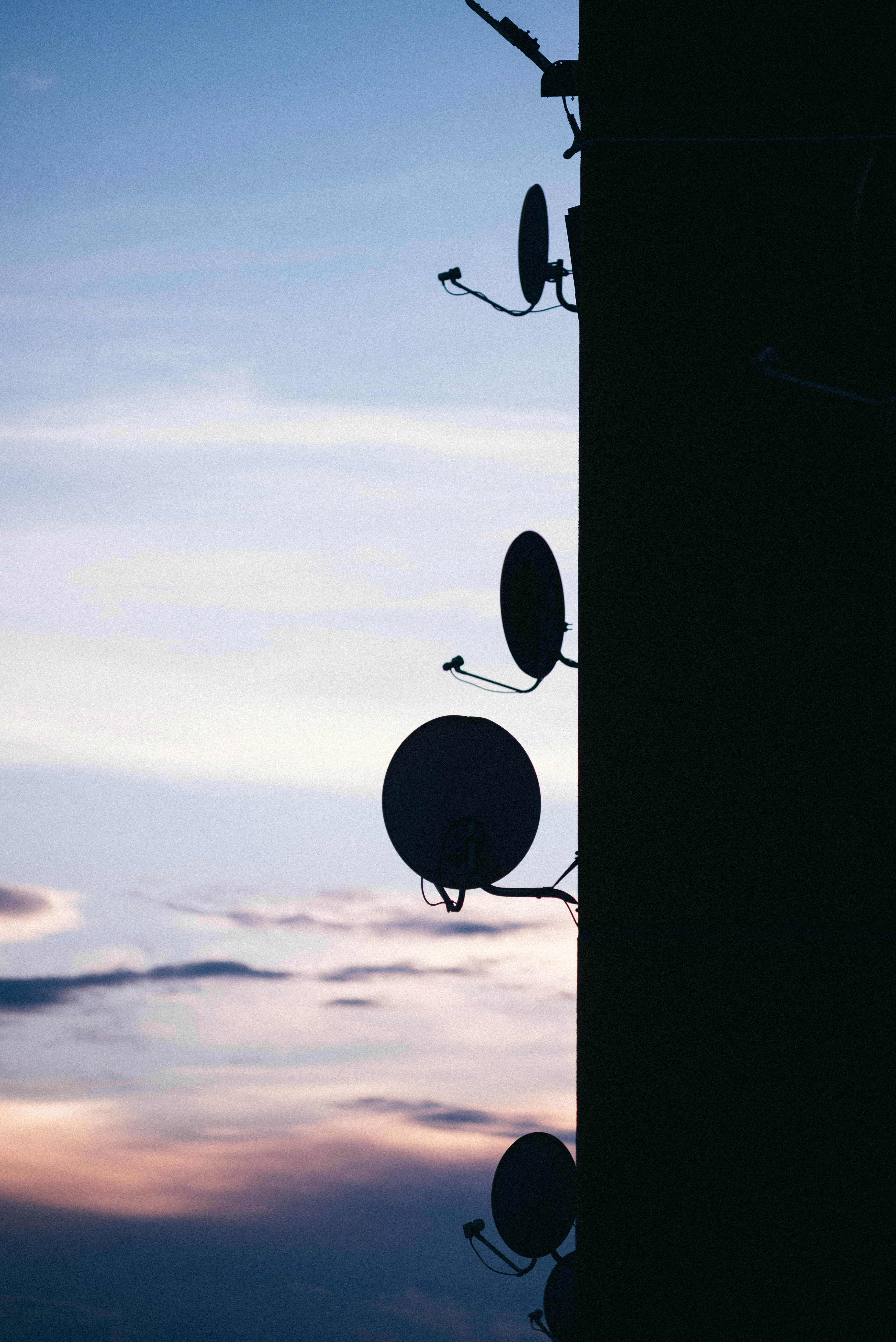 Dusk sky behind a vertical building wall dotted with satellite dishes, forming stark silhouettes against a gradient sunset.