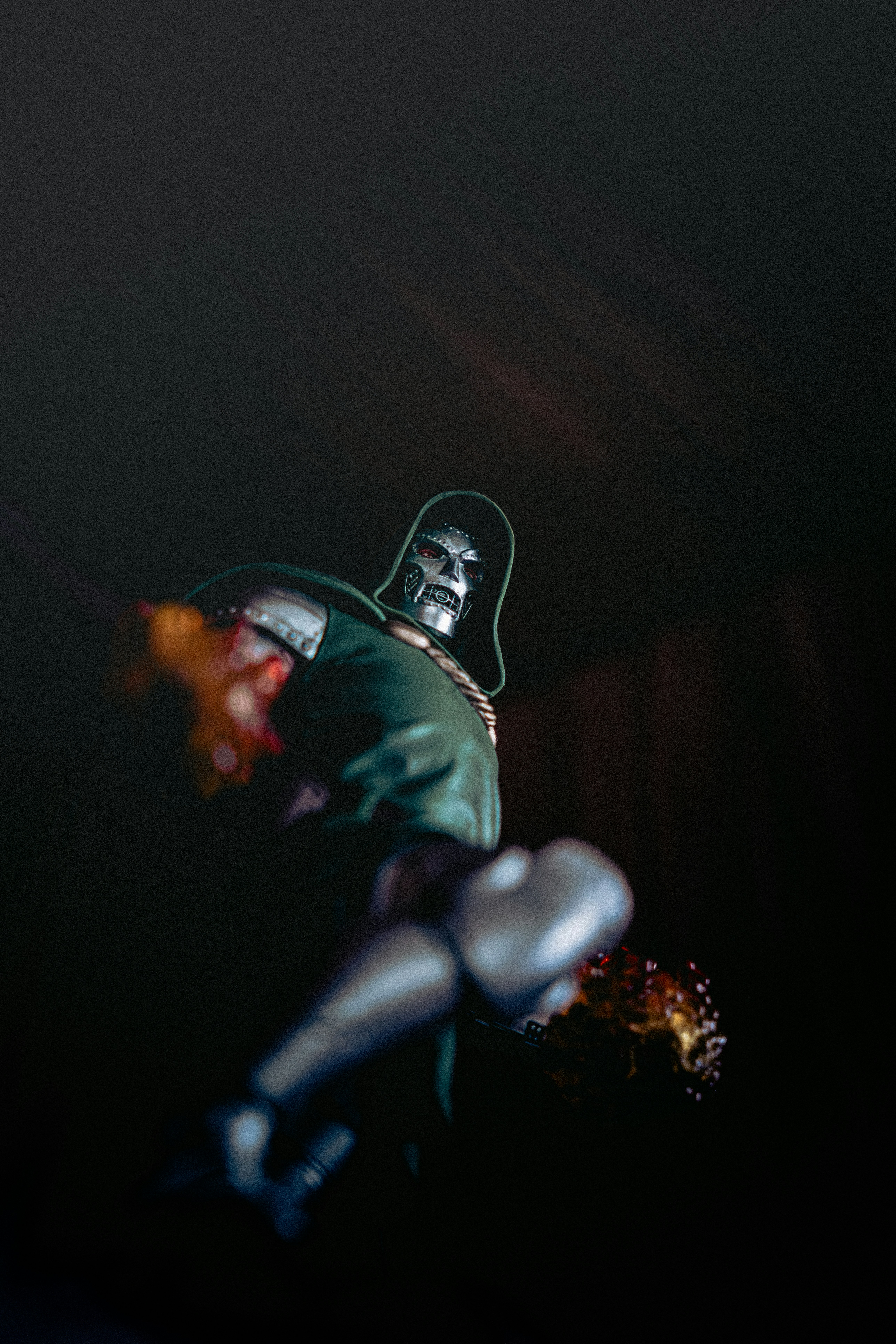A menacing masked figure in a hooded green cloak dominates a dark scene, with a dramatic low angle and a shallow depth of field emphasizing the face and raised arm. A hint of fiery accents adds atmosphere to the composition.