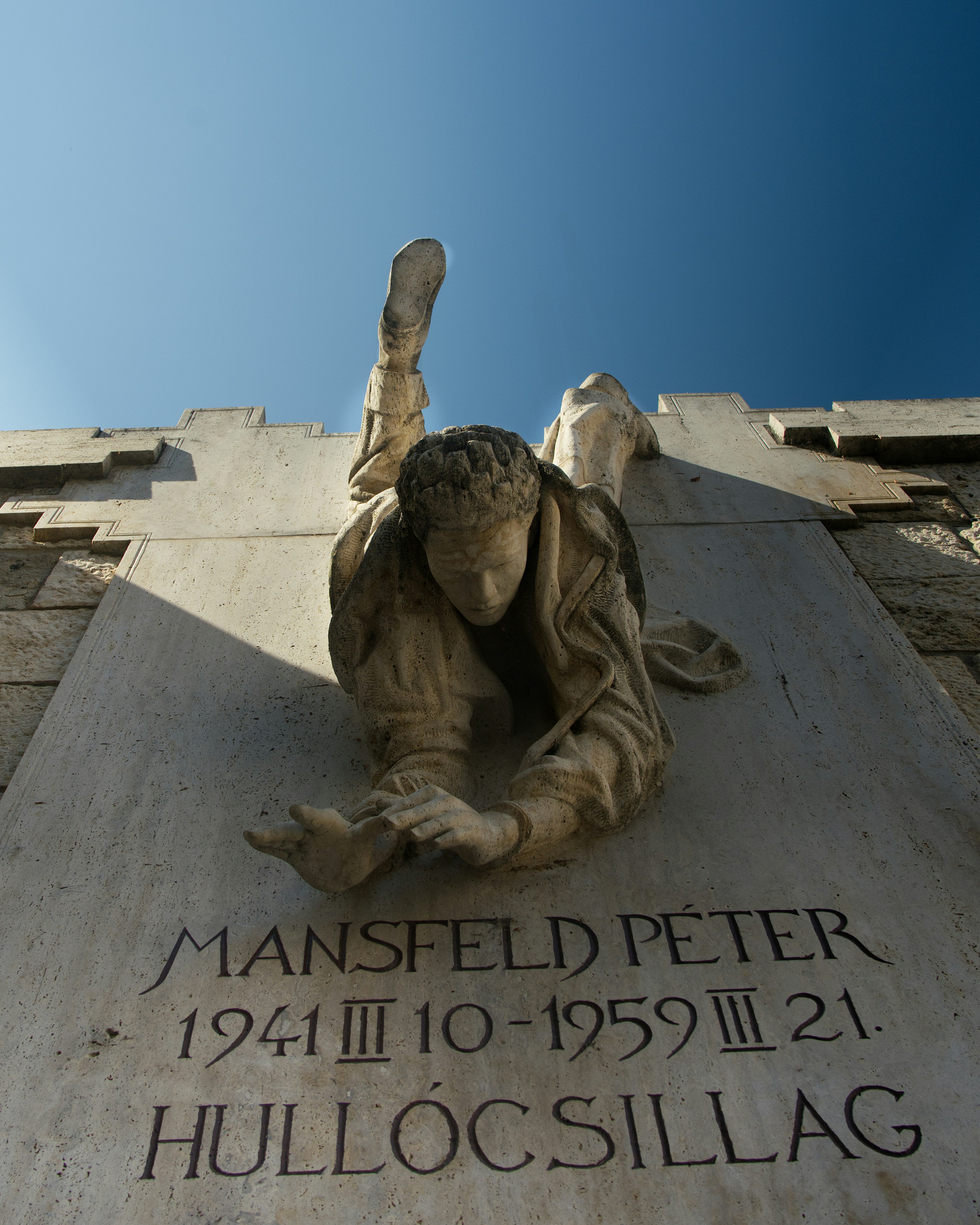 Monumental stone angel relief with inscription beneath, reaching forward as if in motion against a clear blue sky. A solemn memorial sculpture.