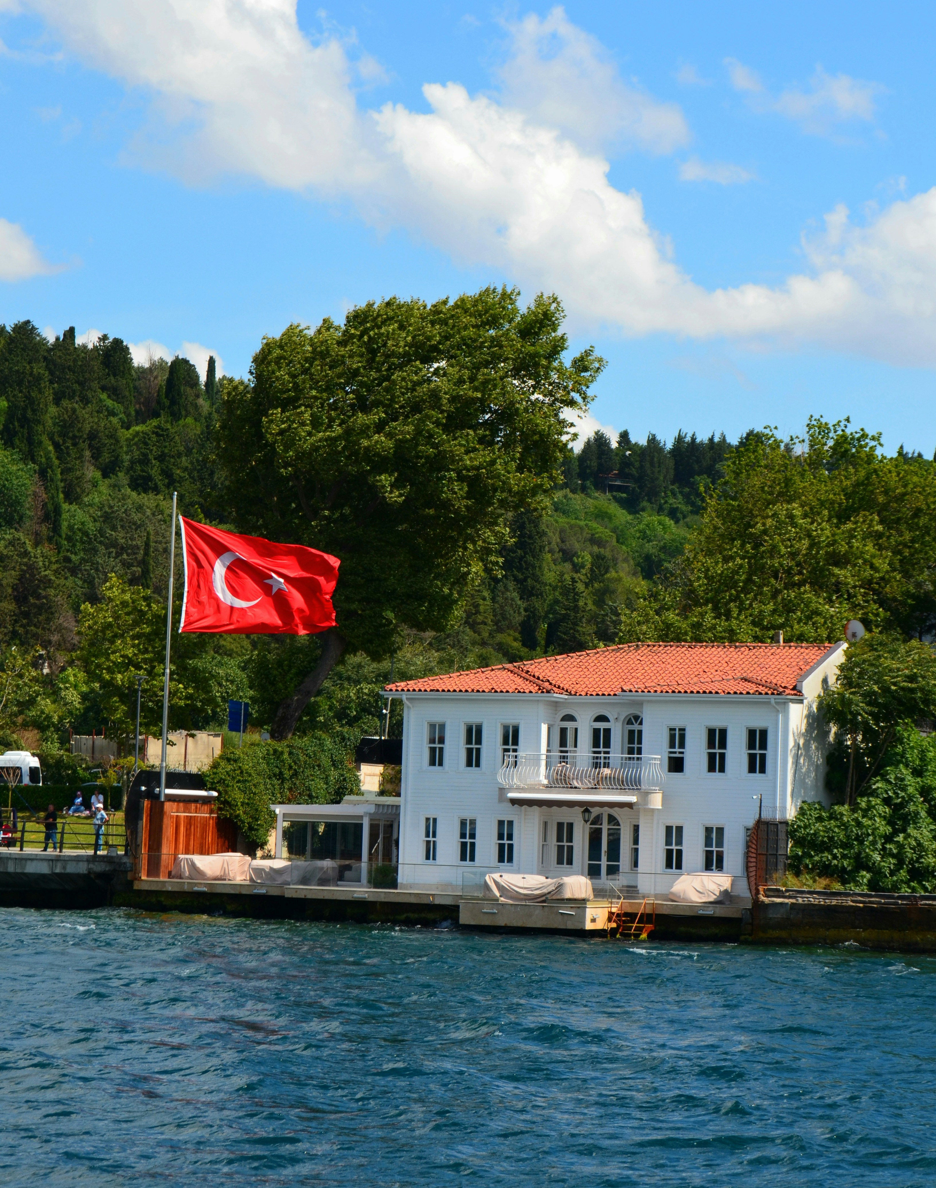 Turkish flag on a pole beside a white lakeside villa with red-tiled roof, calm water in foreground and green hills in the background.