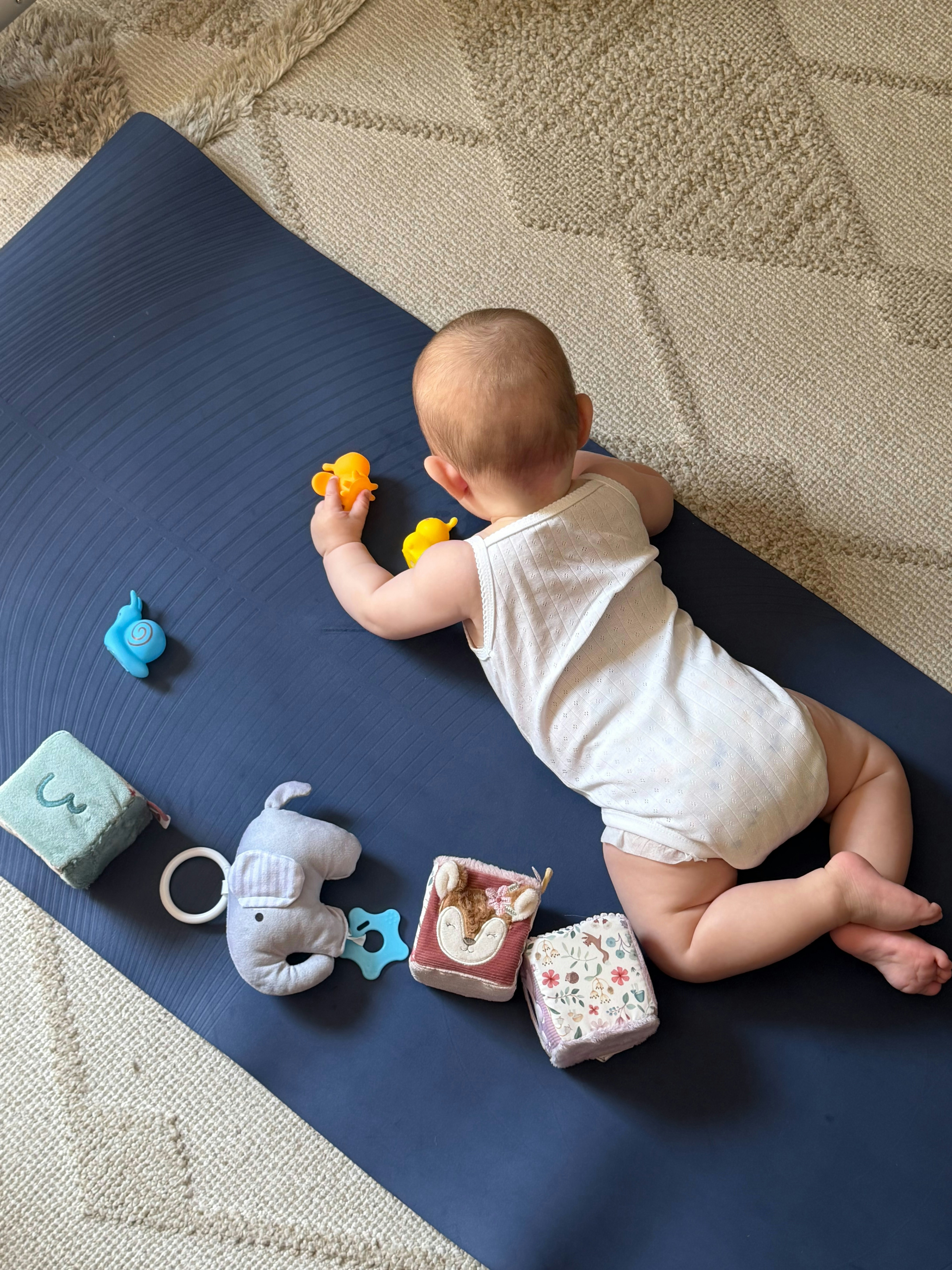 Young child on a dark blue play mat, surrounded by small toys and blocks on a patterned rug.