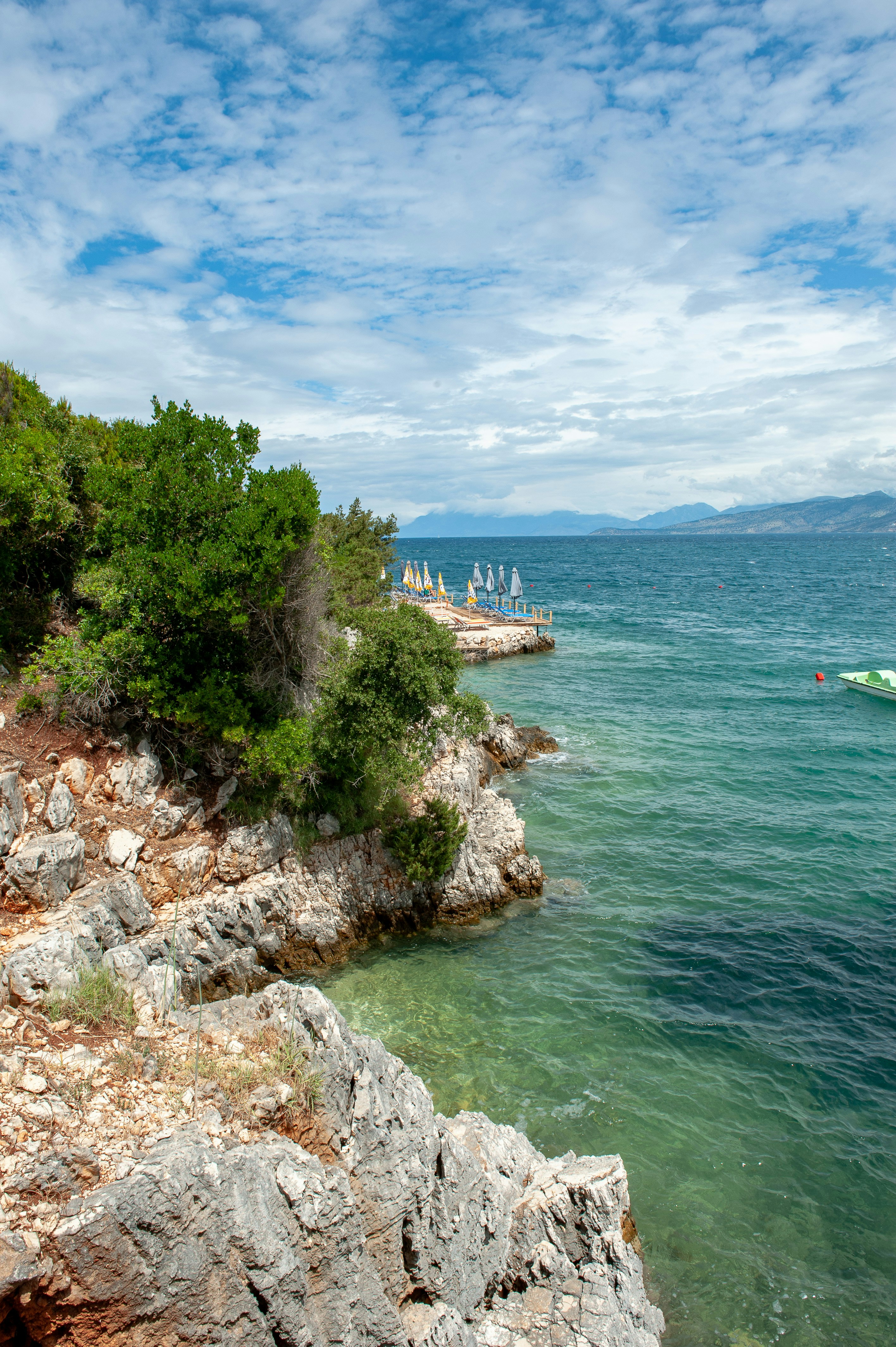 Bright coastal scene with rugged rocky cliffs, green foliage, and clear turquoise water meeting a distant horizon with a small dock and sailboats. A tranquil seaside landscape highlighting natural textures and color.