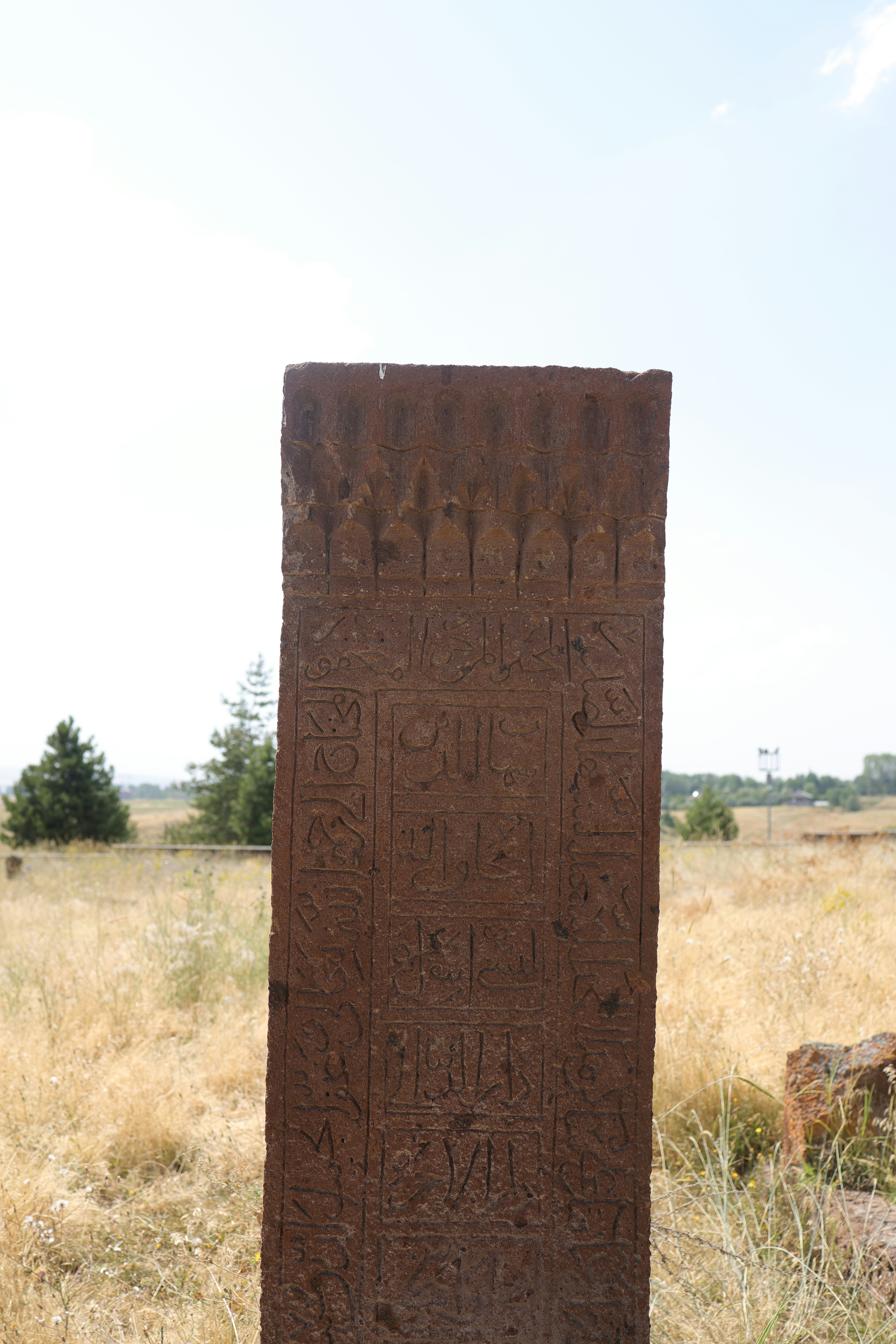 Brown stone obelisk with carved inscriptions stands upright in a dry grassy field under a bright sky.