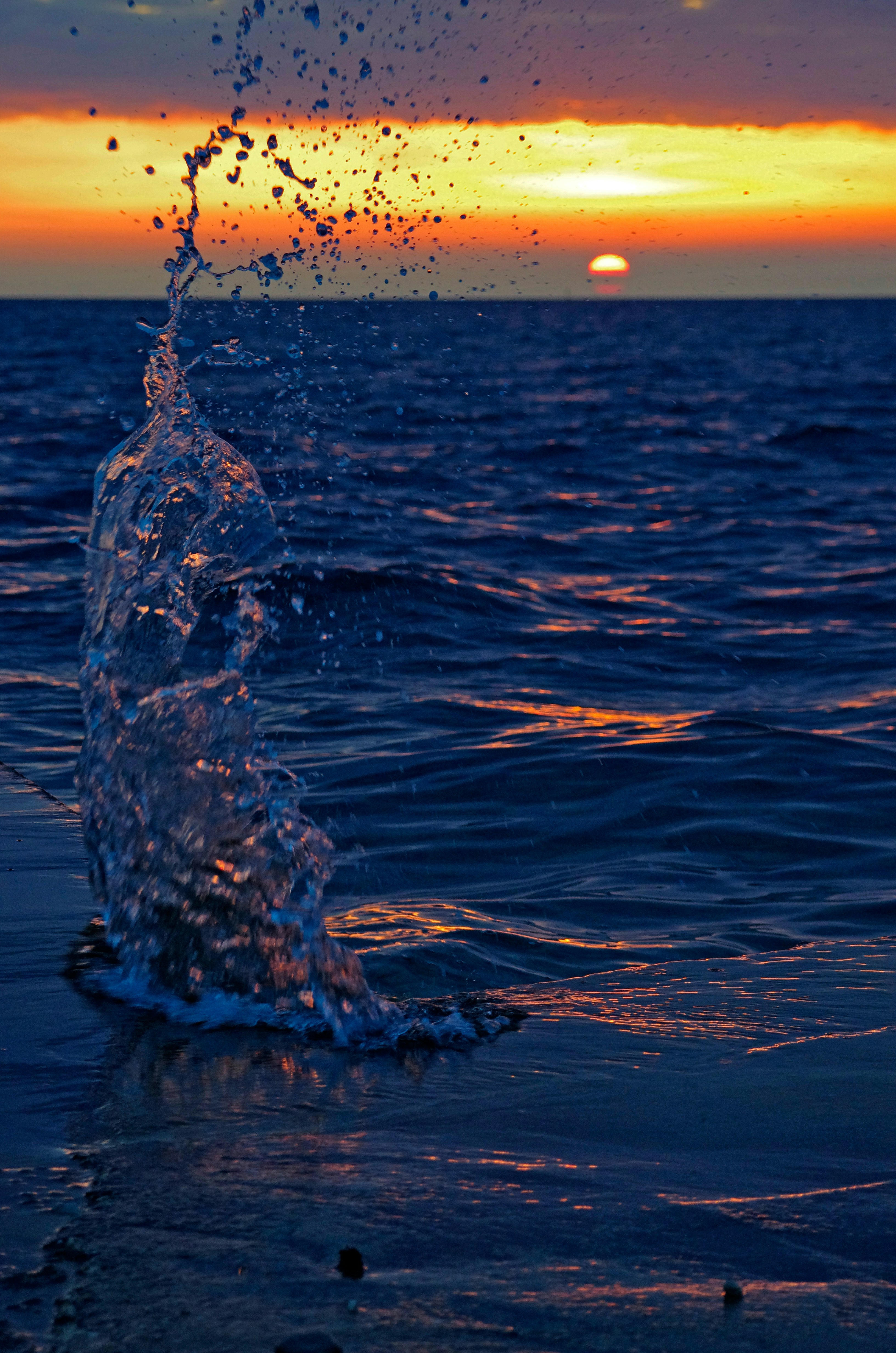 A rising arc of water bursts from the surface during a vibrant sunset over a calm sea, with dramatic blue and orange tones highlighting the spray. The scene captures a single water jet against the horizon.