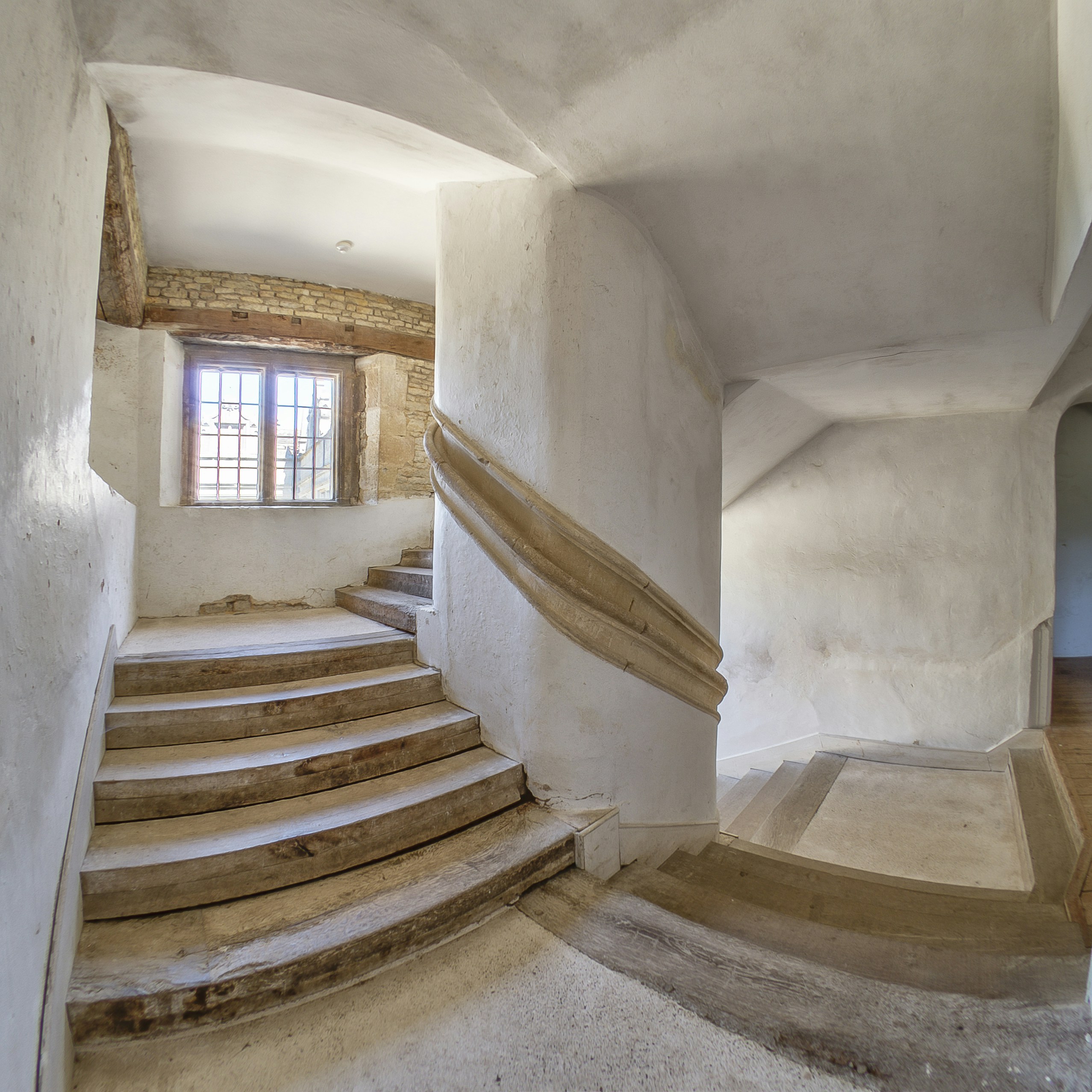 Stone staircase and arching walls bathed in soft daylight from a small window; a medieval interior with curved banister and plaster textures.