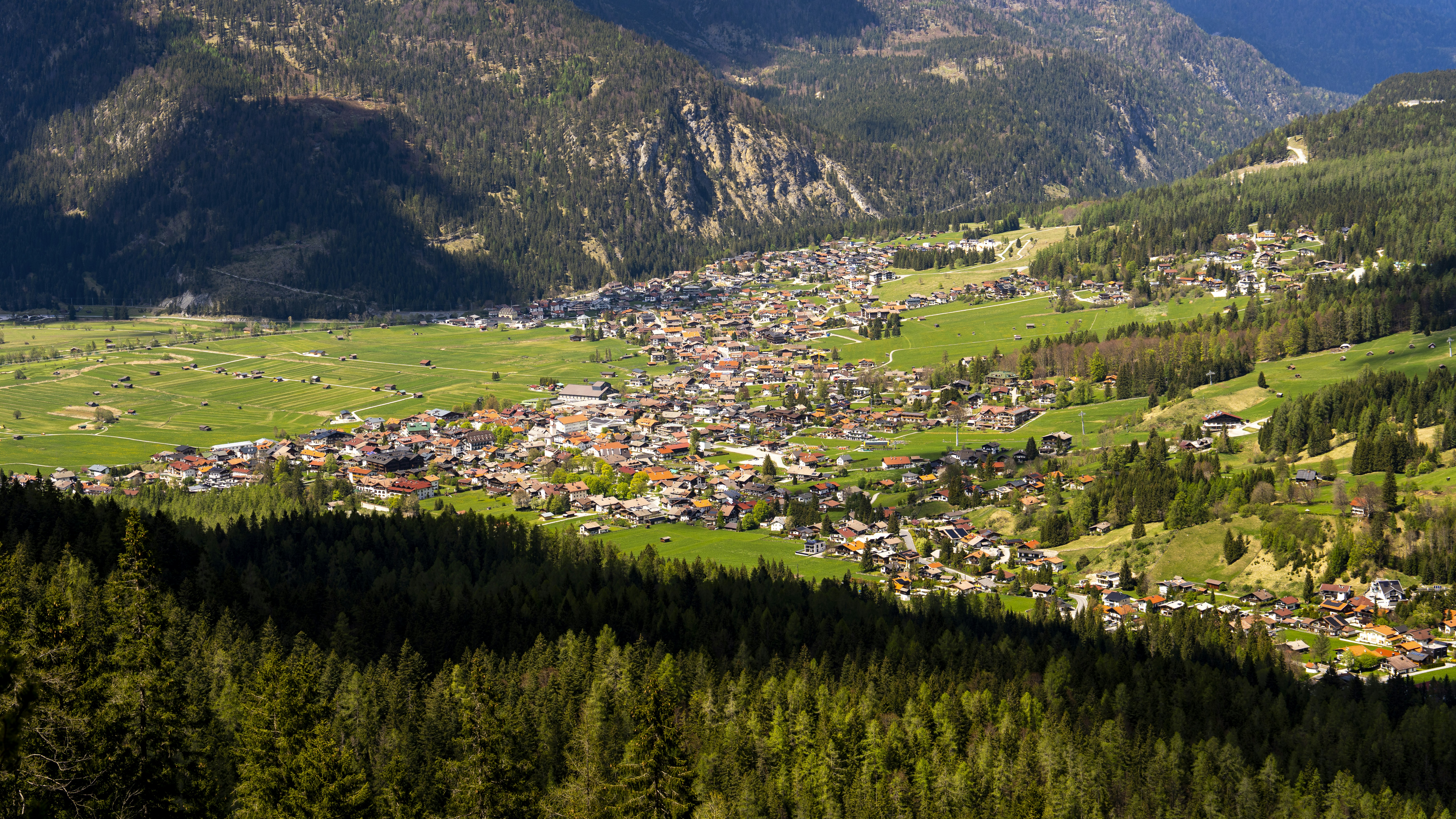 Colorful village nestled in a green valley with surrounding forest and distant mountains; wide landscape showing cultivated fields and climbing roads.