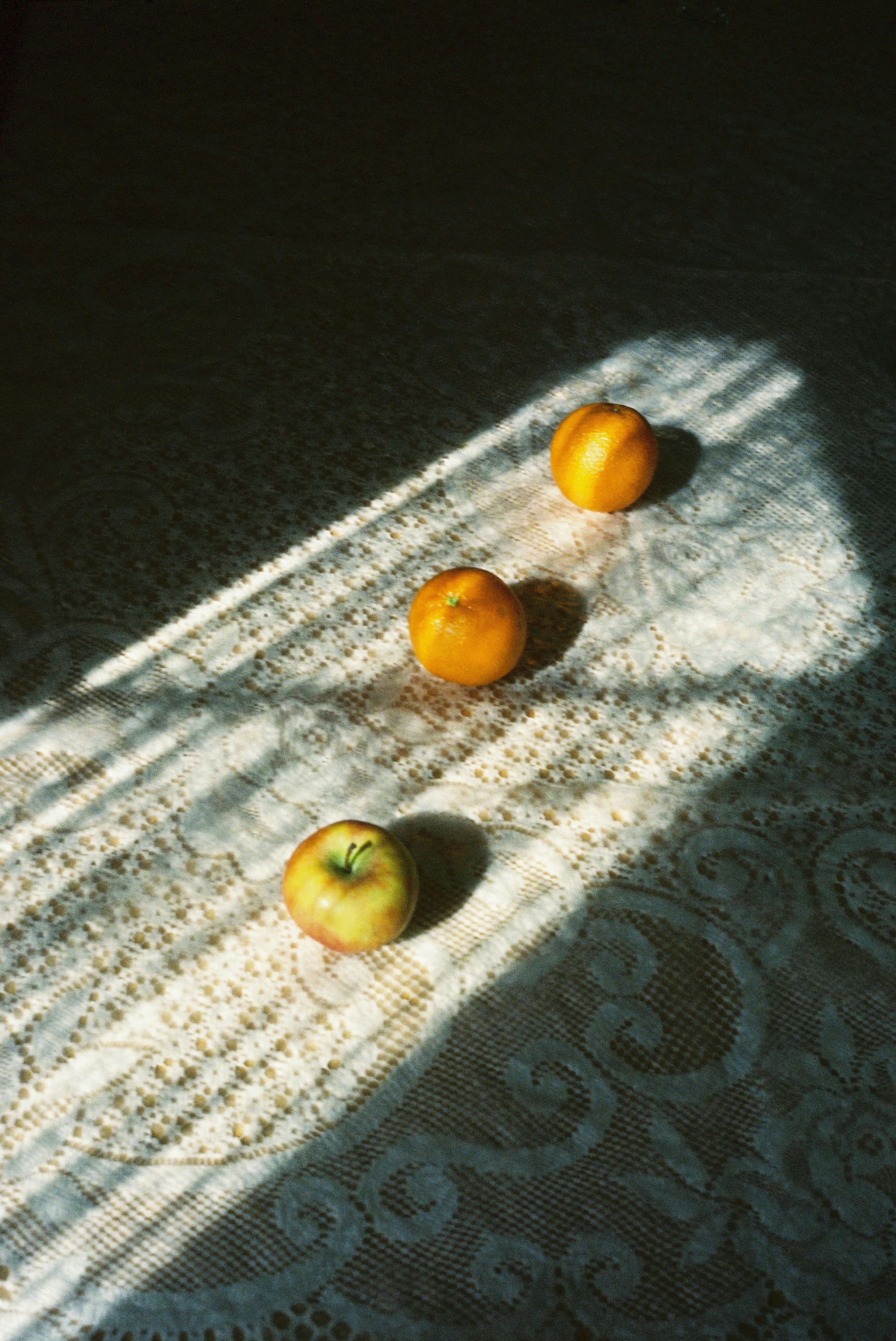 Three fruit pieces bathed in diagonal sunlight on a textured lace surface, casting long shadows and highlighting warm tones.
