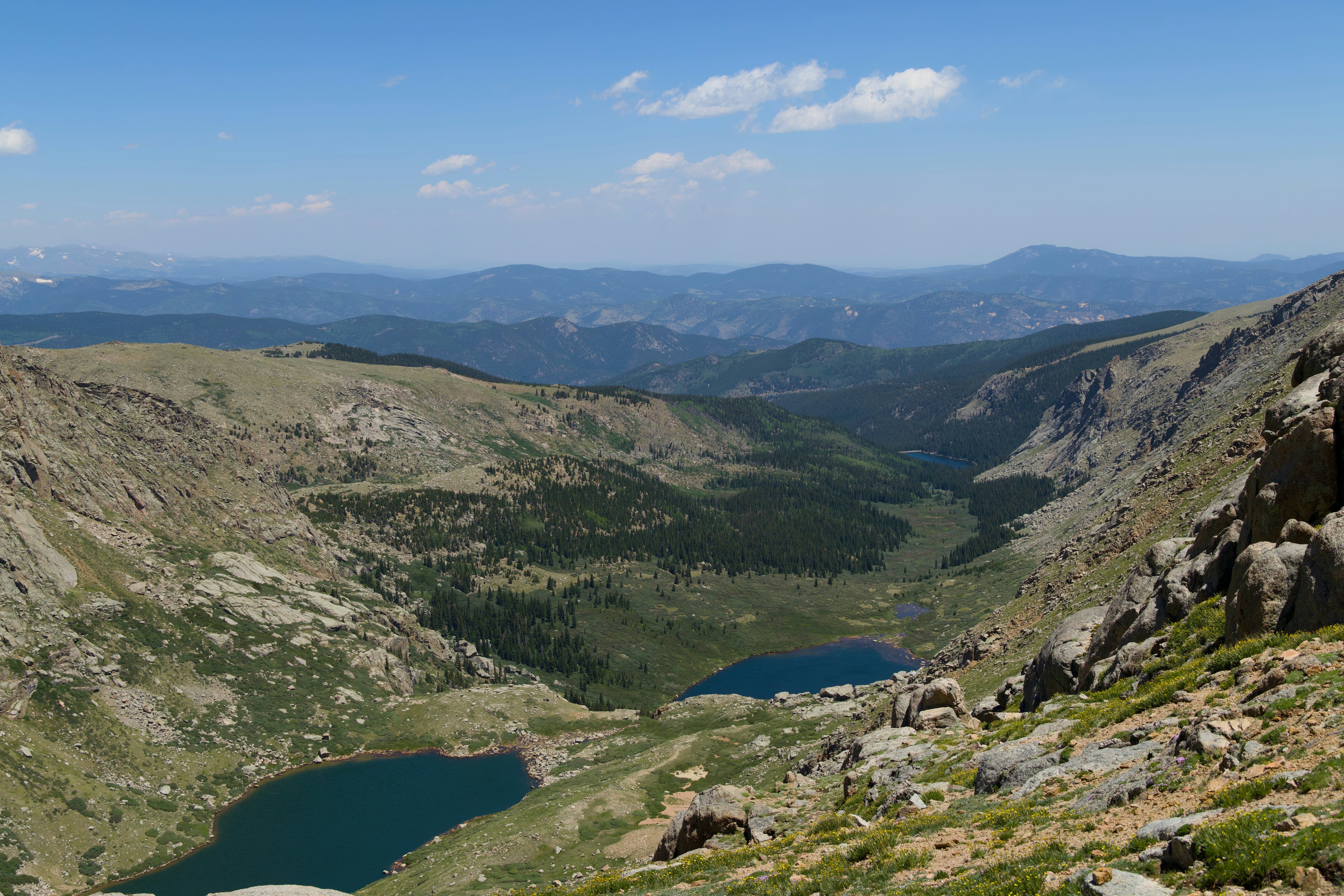 Mountain valley with emerald lakes and rocky slopes under a bright blue sky. Aerial perspective reveals rugged terrain and distant ridgelines, highlighting the scale of the alpine basin.